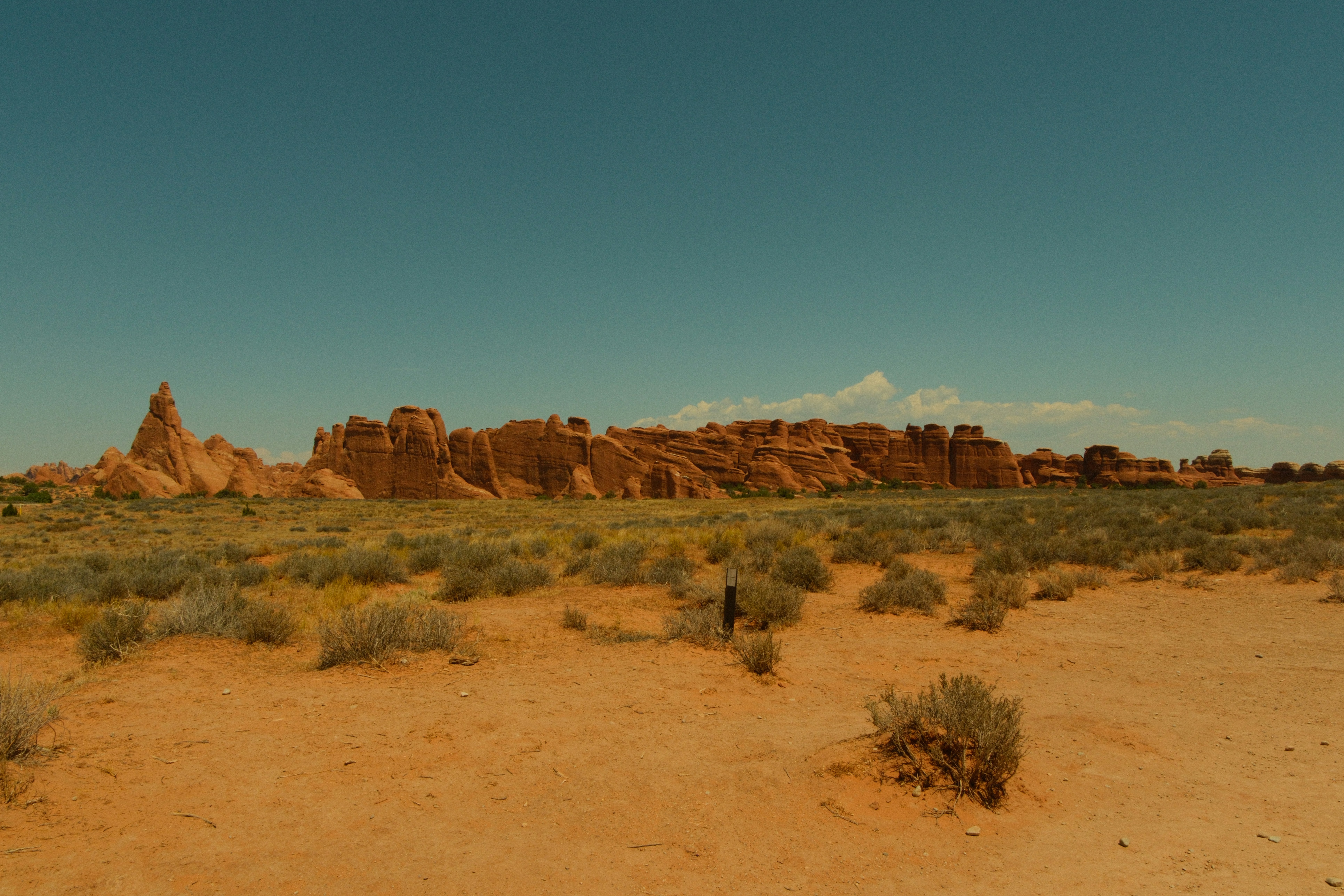 Desert landscape with eroded rock formations on the horizon and sparse scrub in the foreground; warm canyon hues under a clear blue sky. Identifiable as a photographic landscape, not a painting or illustration.
