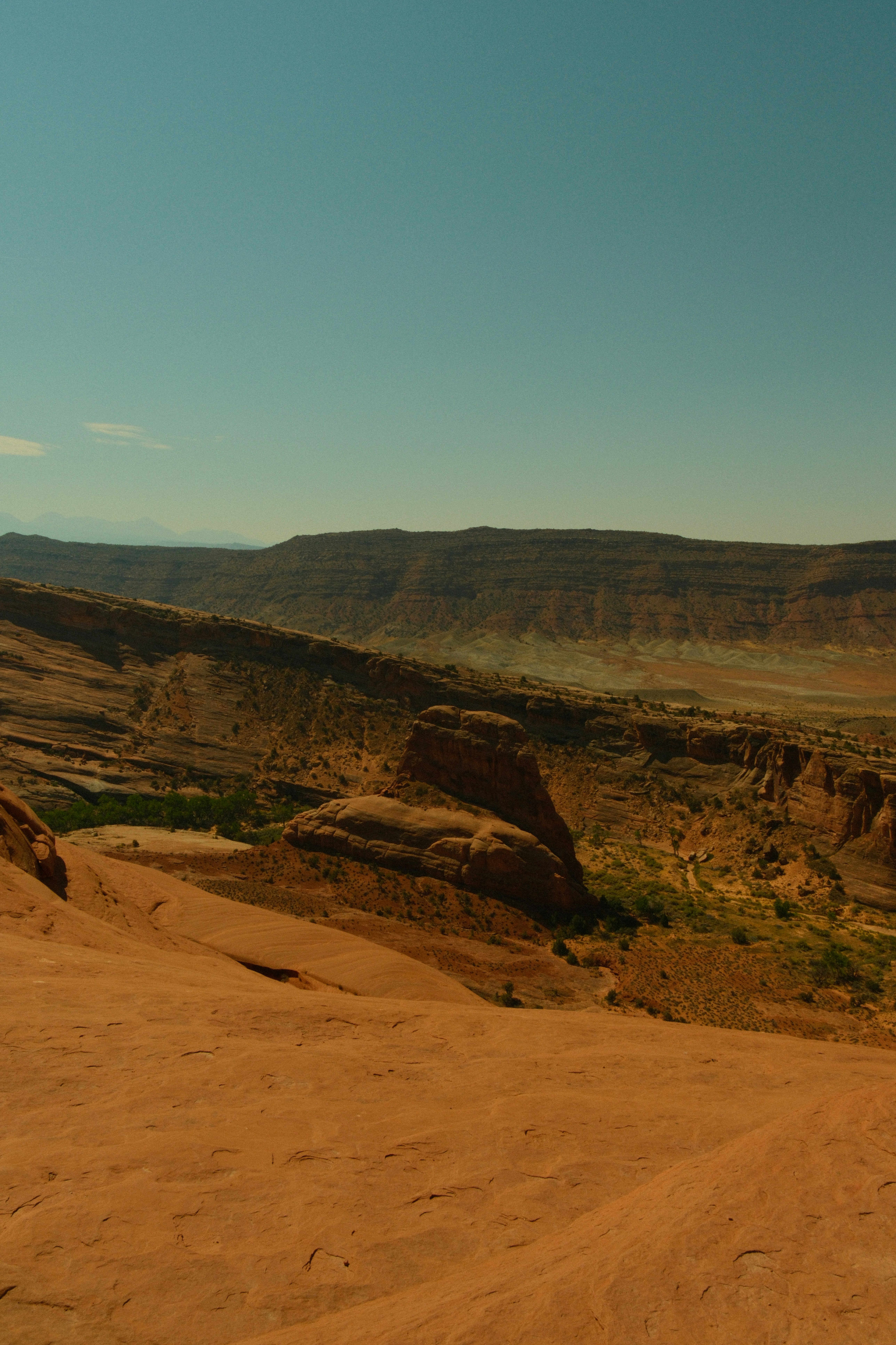 Rugged red rock formations stretch across a desert plateau with a pale blue sky above, capturing a wide, sunlit landscape.
