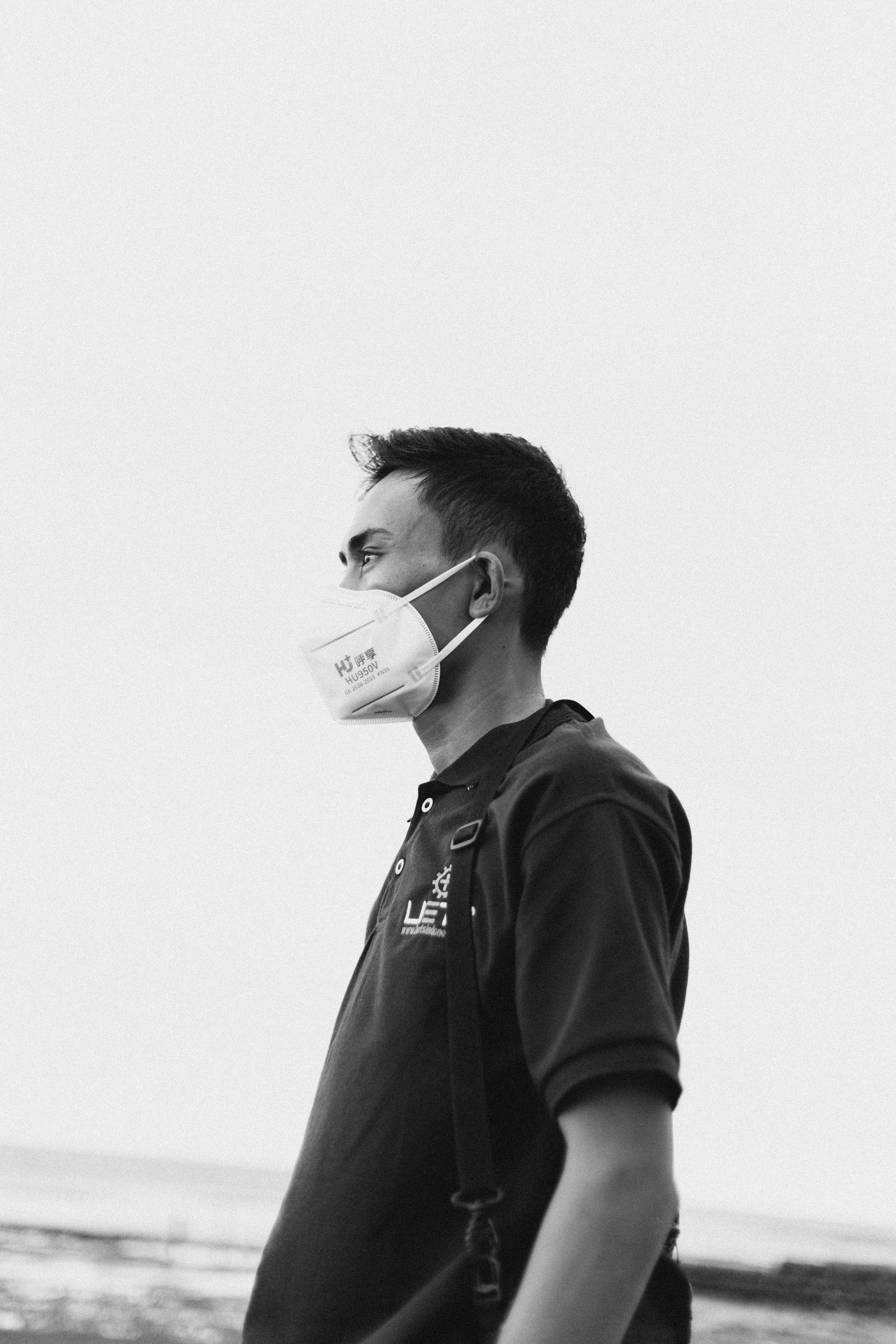 A man wearing a protective mask stands in profile against a pale, minimalist backdrop near a shoreline.