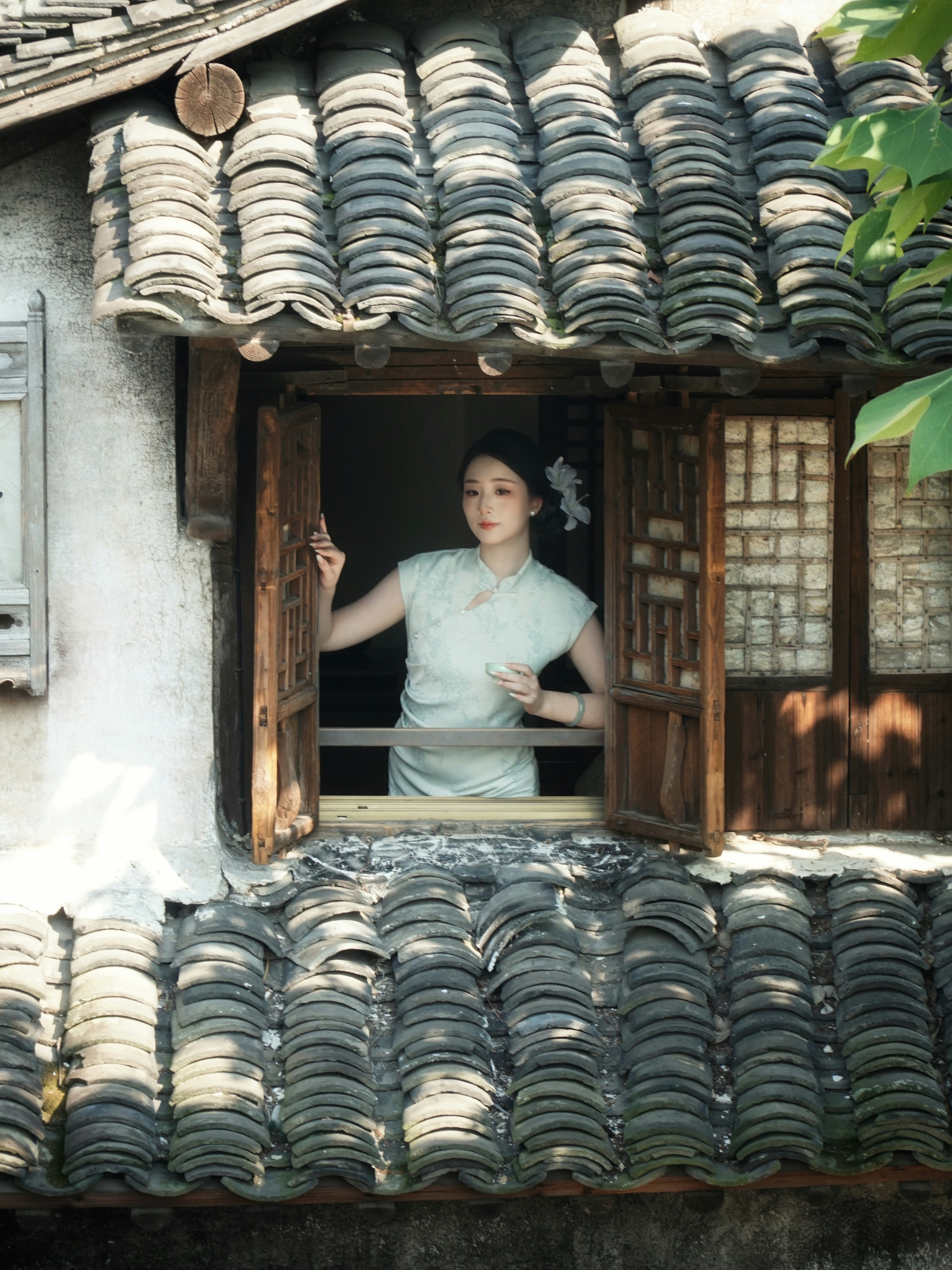 A woman in a qipao stands at a rustic-window opening, gazing outward with a calm, composed expression. The scene is framed by weathered roof tiles and wooden lattice, suggesting a blend of heritage and daily life.