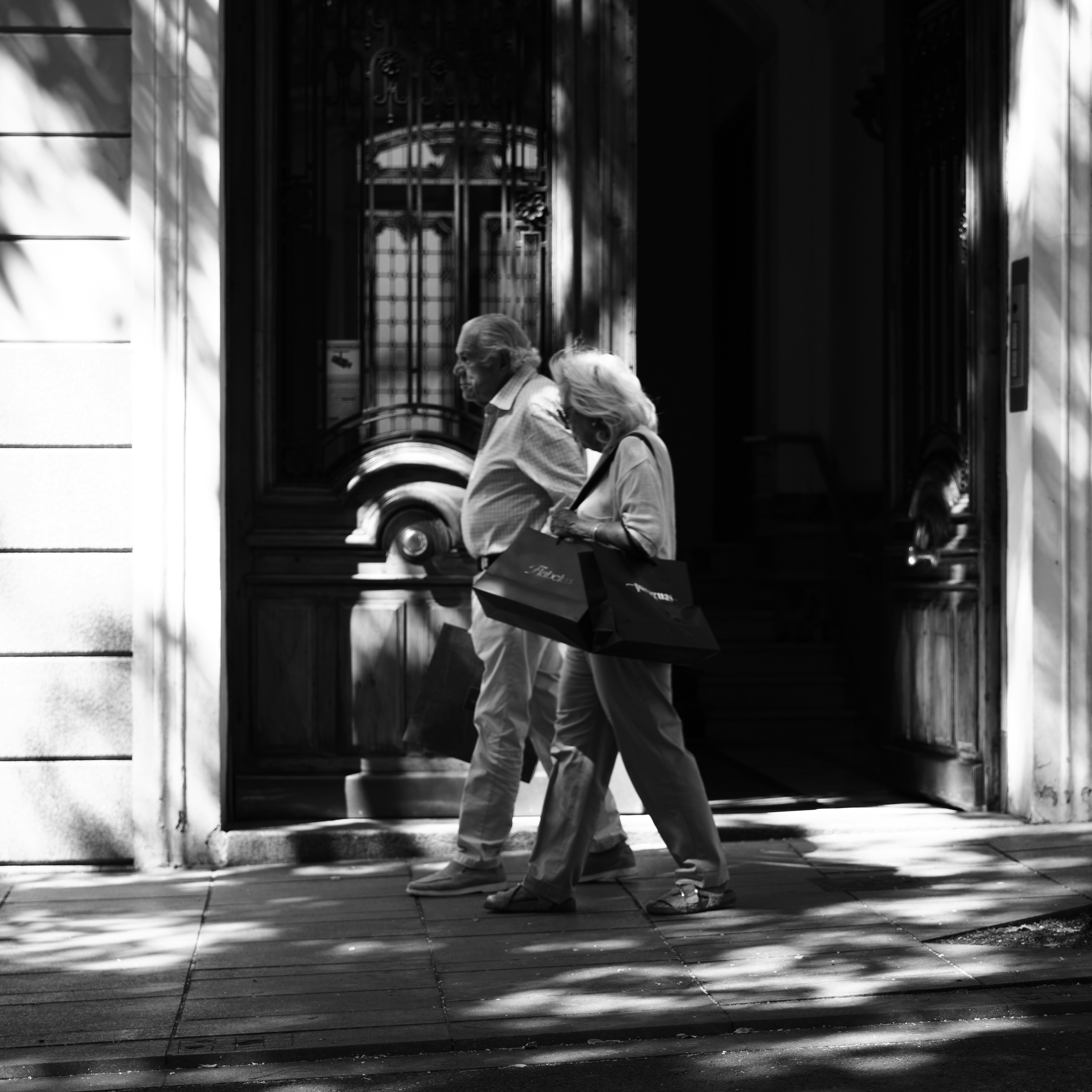 Walking in Shade: Elder Couple Crossing a Sun-Dappled City Threshold