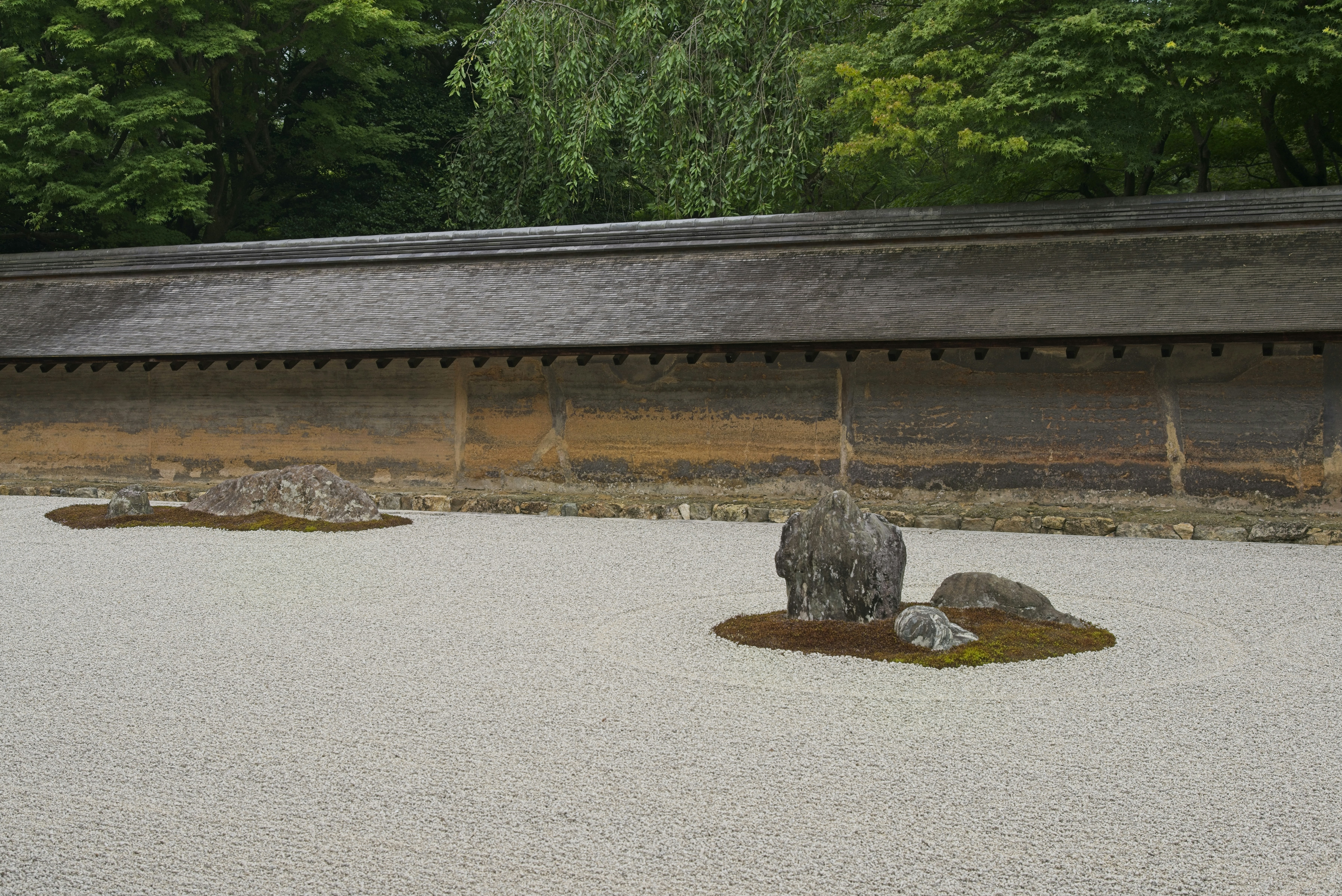 The world-famous rock garden of Ryōan-ji in Kyoto displays its minimalist stone groupings, with weathered boulders resting on moss islands amid meticulously raked white gravel, on a hot, hazy summer afternoon in July. A long, weathered earthen wall streaked with age borders the garden beneath a wooden tiled roof, while dense willow and maple trees rise above it. The sparse, deliberate arrangement of stone and gravel embodies the essence of Zen dry landscape design, offering a quiet, timeless space for contemplation within Kyoto's historic temple grounds.