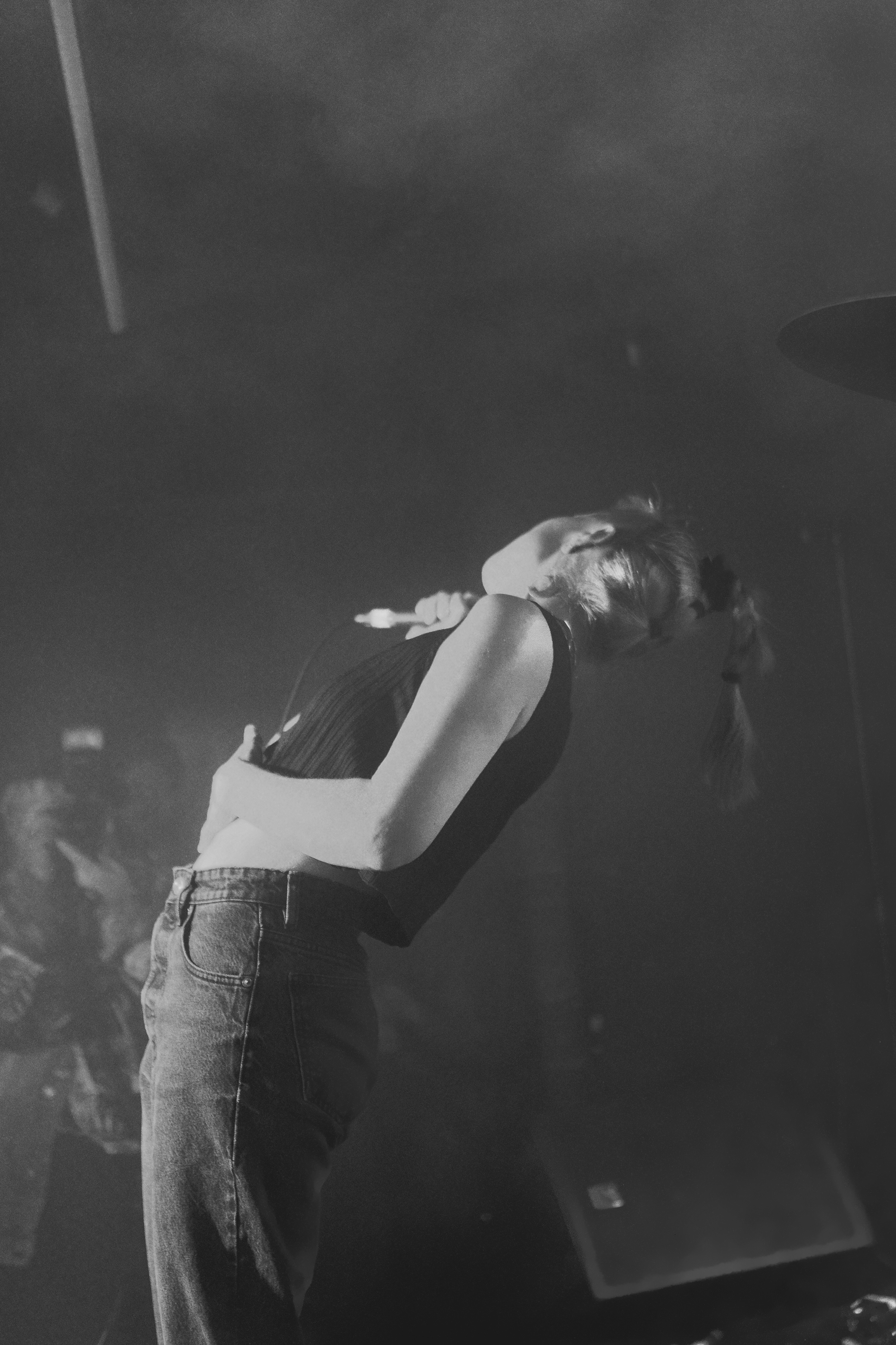 A grayscale live-performance shot capturing a vocalist mid-lean, microphone gripped, with smoke and audience silhouettes in the hazy background. The scene emphasizes energy and motion on stage.