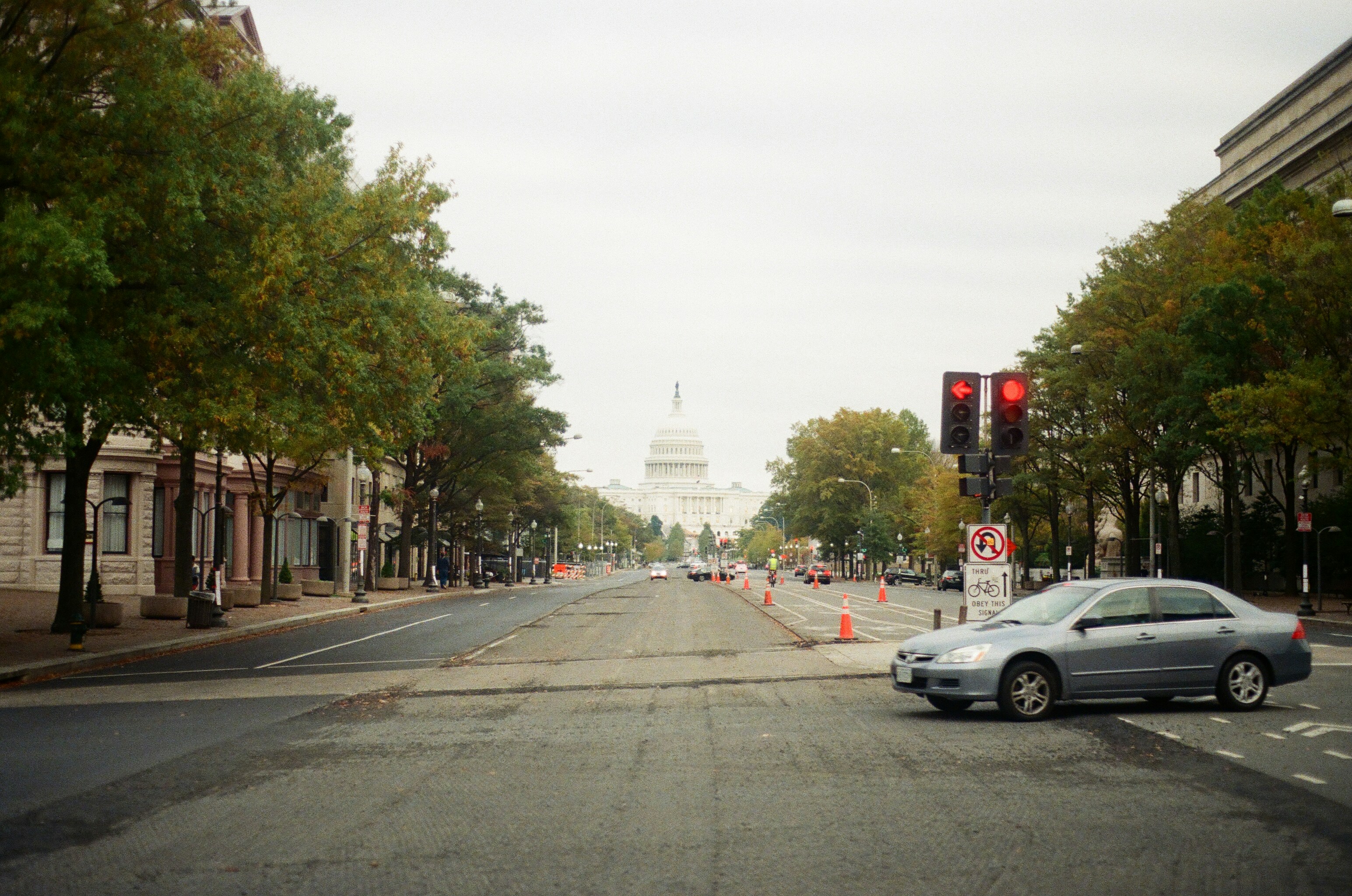 City street scene with a distant capitol dome, traffic lights, and a lone car in the foreground; composed as a linear perspective toward the distant government building. A subtle, wide-angle view of urban life with autumn trees lining the roadway.