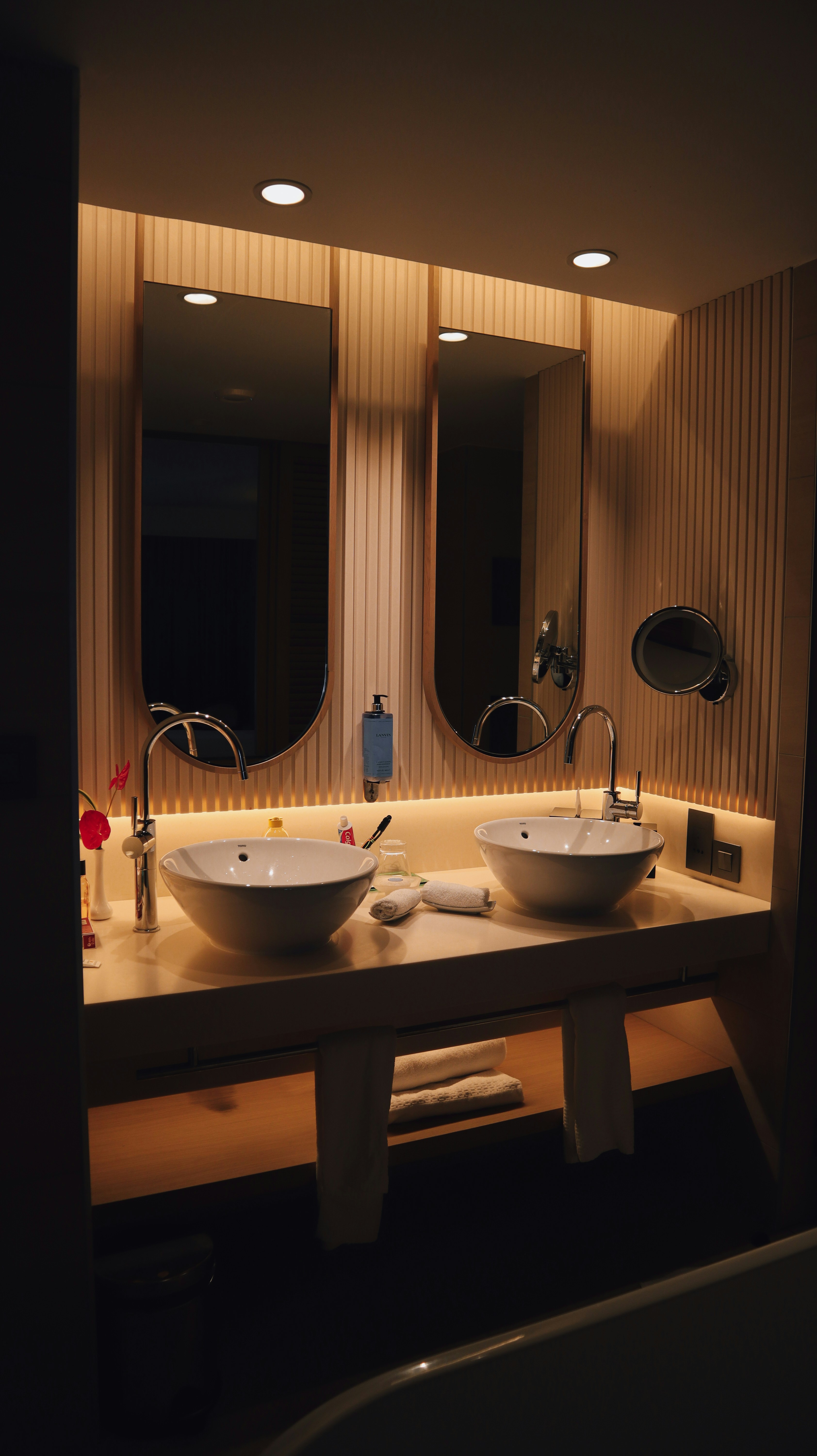 Two vessel sinks sit under warm lighting in a modern bathroom vanity with mirrors and a round magnifying mirror. A clean countertop holds toiletries while towels hang beneath the counter.