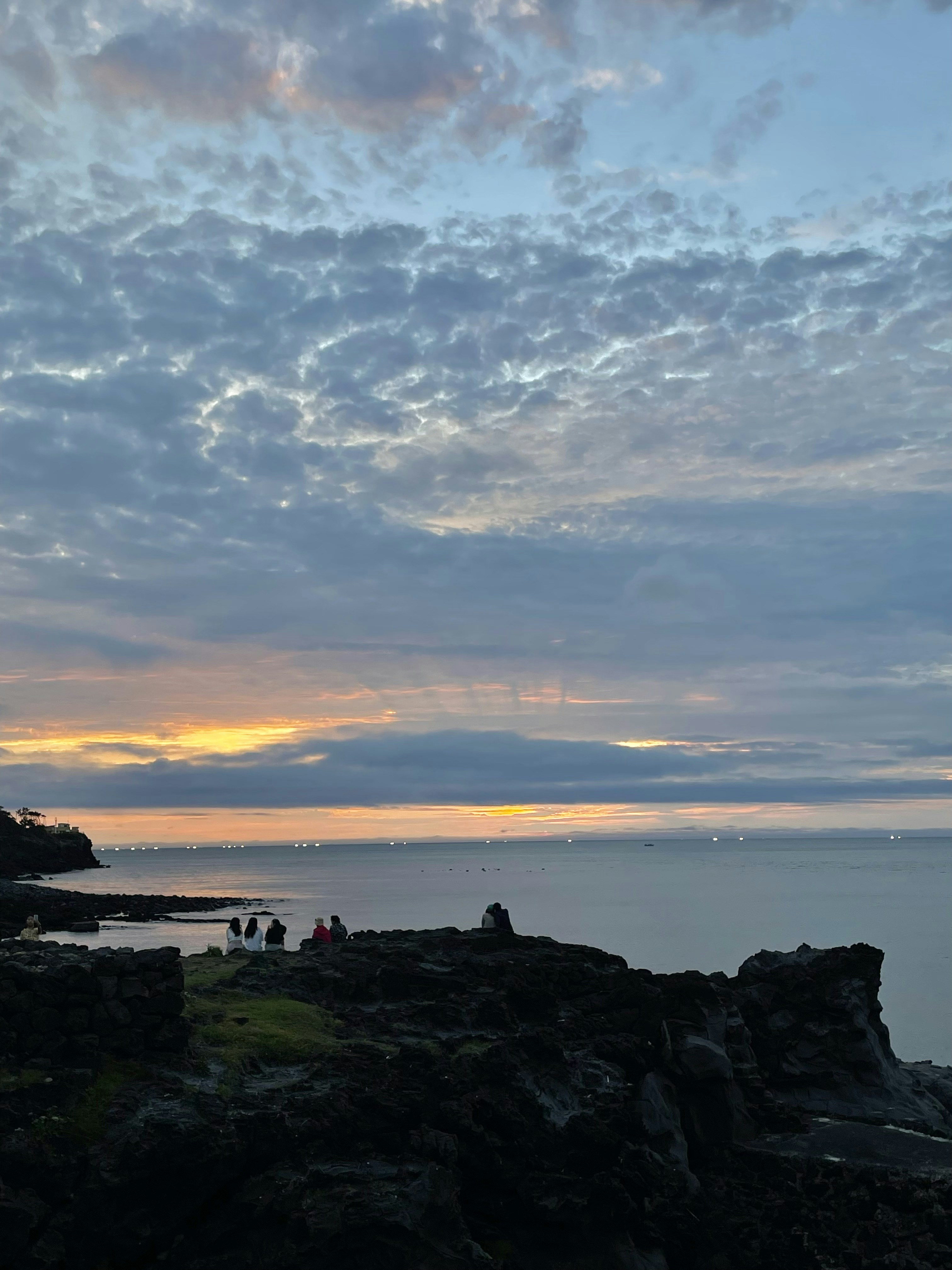 Sunset over calm water with silhouettes of people on rocky shoreline and a cloudy sky. A tranquil coastal scene highlighting the horizon and soft colors.