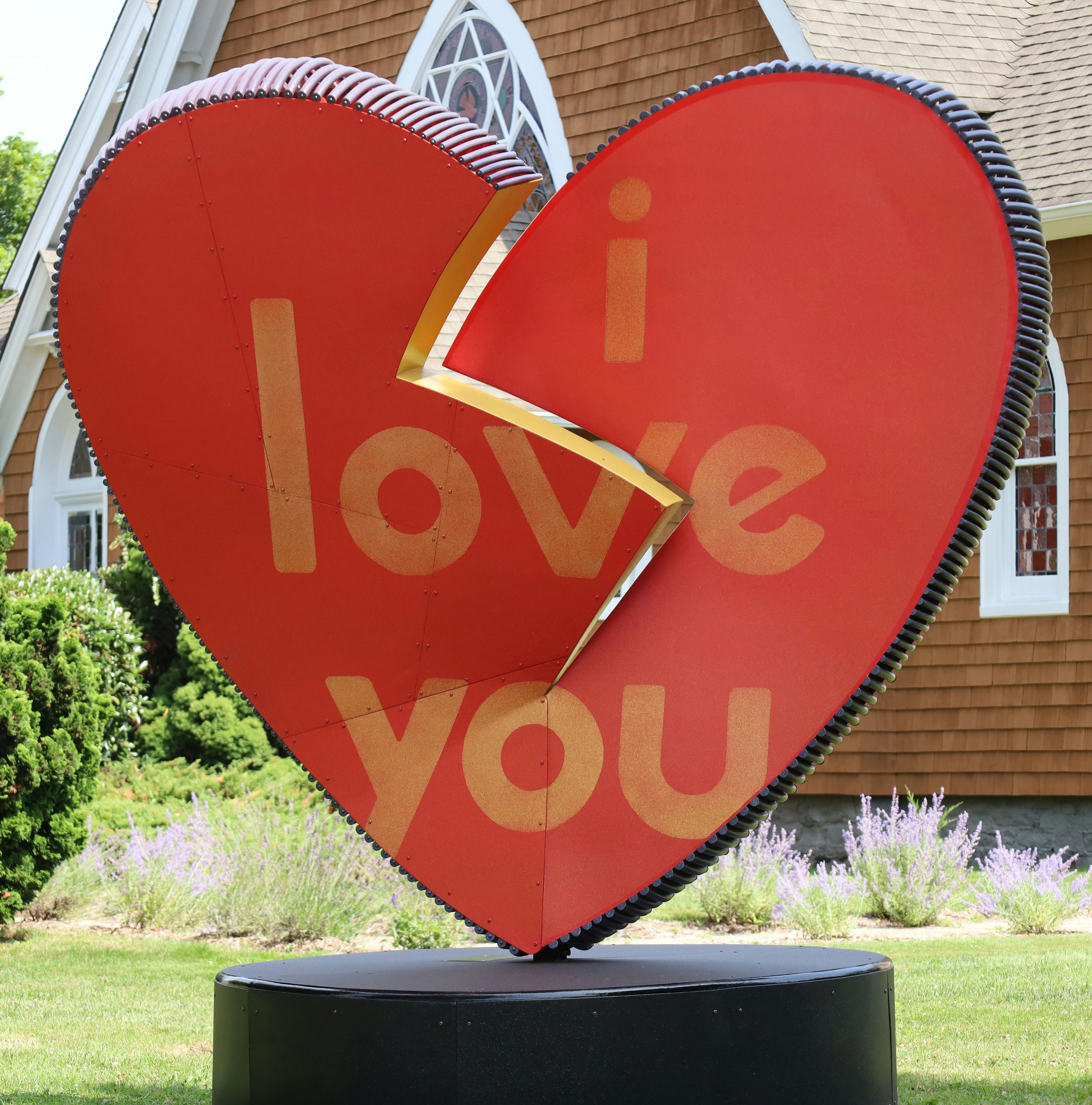 A vivid red broken-heart sculpture with the words 'love you' is displayed outdoors beside a house, mounted on a black pedestal amid green landscaping.