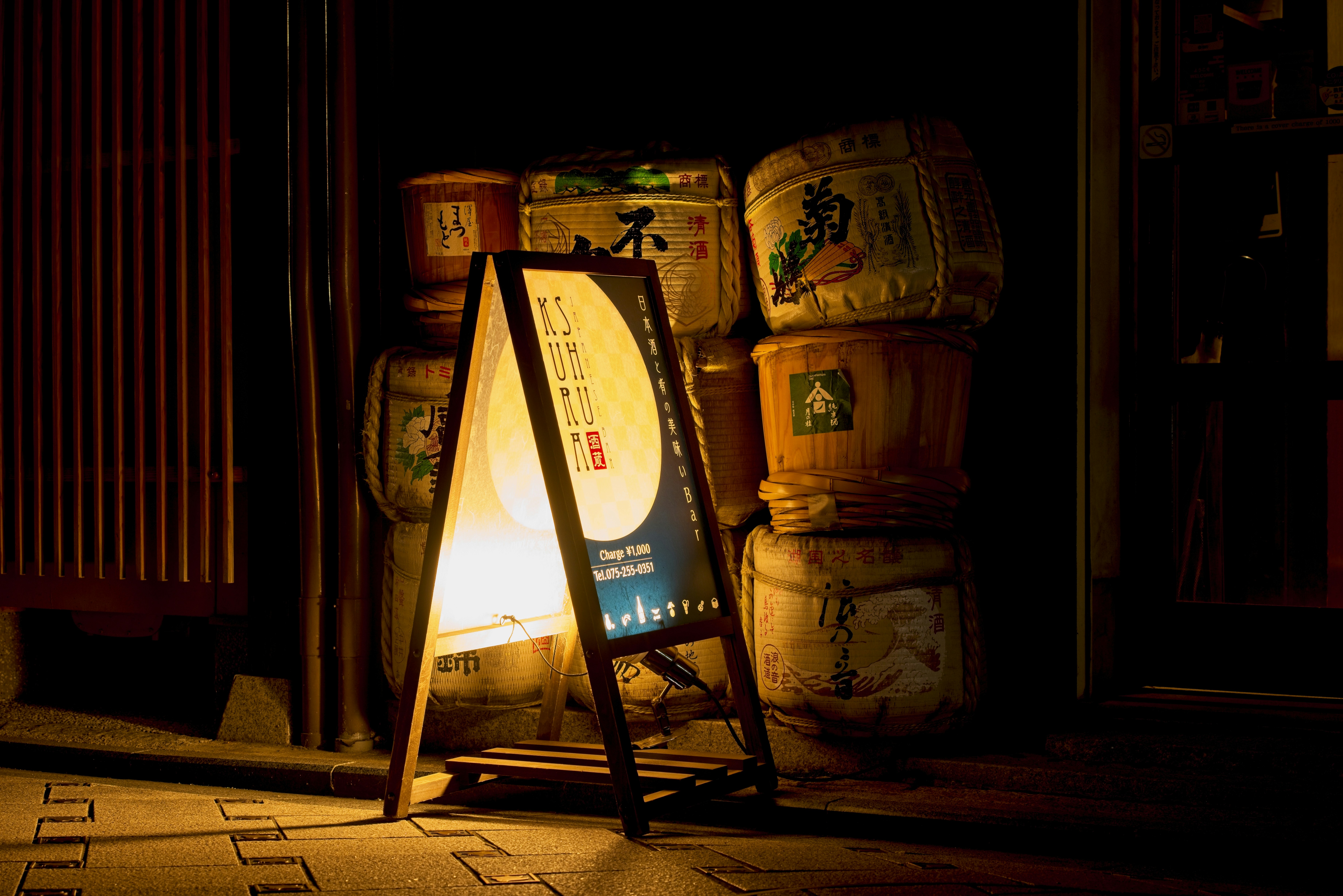 A glowing A-frame signboard for a Japanese sake bar illuminates the entrance to an establishment near Pontocho Alley, Kashiwayachō, Kyoto, on a humid July evening. Behind it, a stack of decorative straw-wrapped sake barrels bearing bold kanji labels adds authentic character to the scene. The warm backlit sign advertises the bar's charge and contact details, creating an inviting glow against the dark alley, a quintessential glimpse of Kyoto's traditional sake culture after dark.