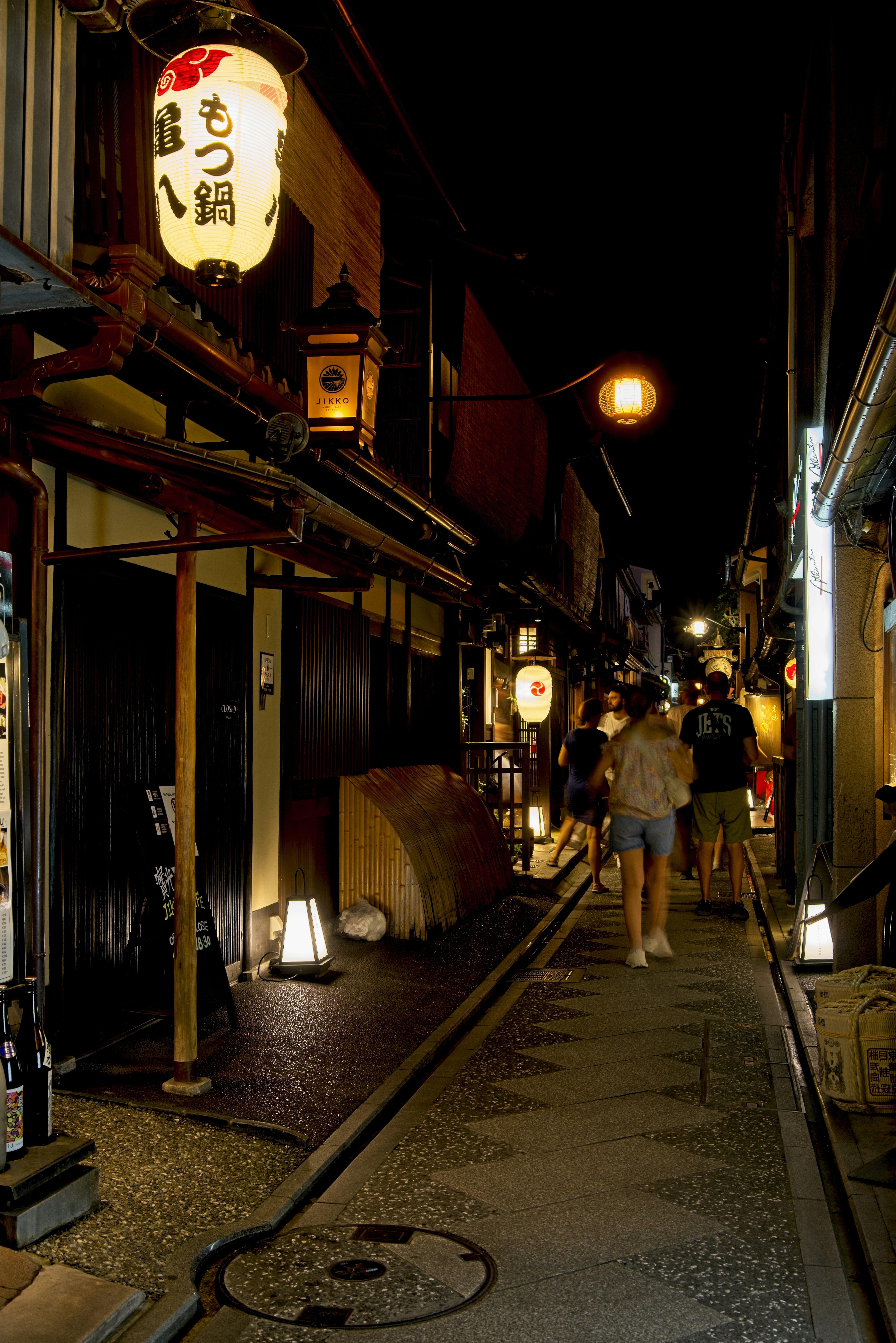 A large glowing lantern advertising motsunabe hangs above a traditional restaurant entrance along Pontocho Alley, Kashiwayachō, Kyoto, as tourists stroll the narrow stone lane on a warm July evening. Round paper lanterns and wooden signboards illuminate the historic wooden facades lining both sides, while bamboo fencing and stacked sake crates add authentic texture. The lively yet intimate atmosphere captures Kyoto's classic nighttime dining district in full swing.