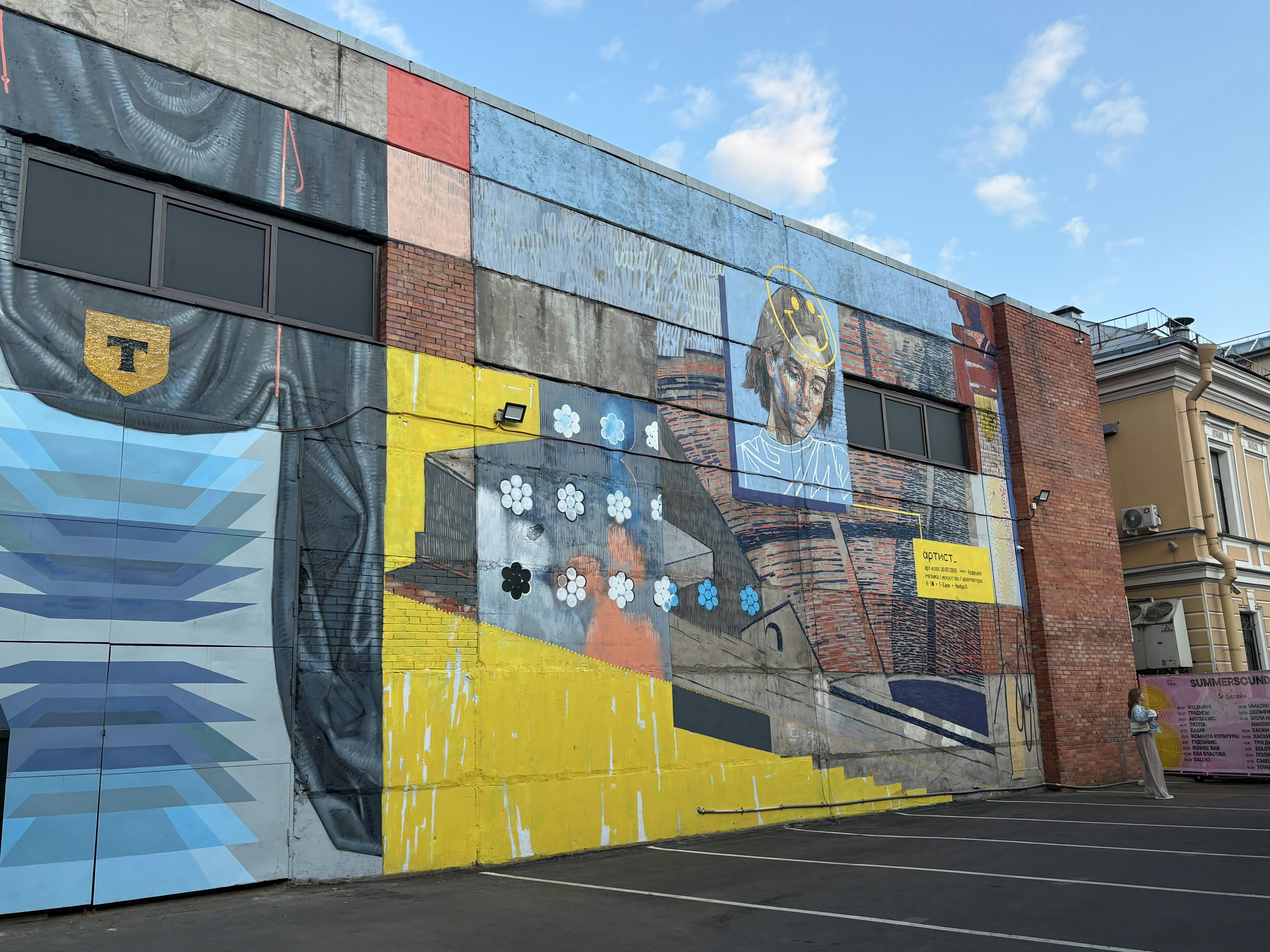 Vibrant mural on an urban brick wall featuring geometric patterns, a sunken yellow stairway, and a stylized portrait at the upper center. The artwork fills a warehouse-like facade, with neighboring building facades visible to the right.
