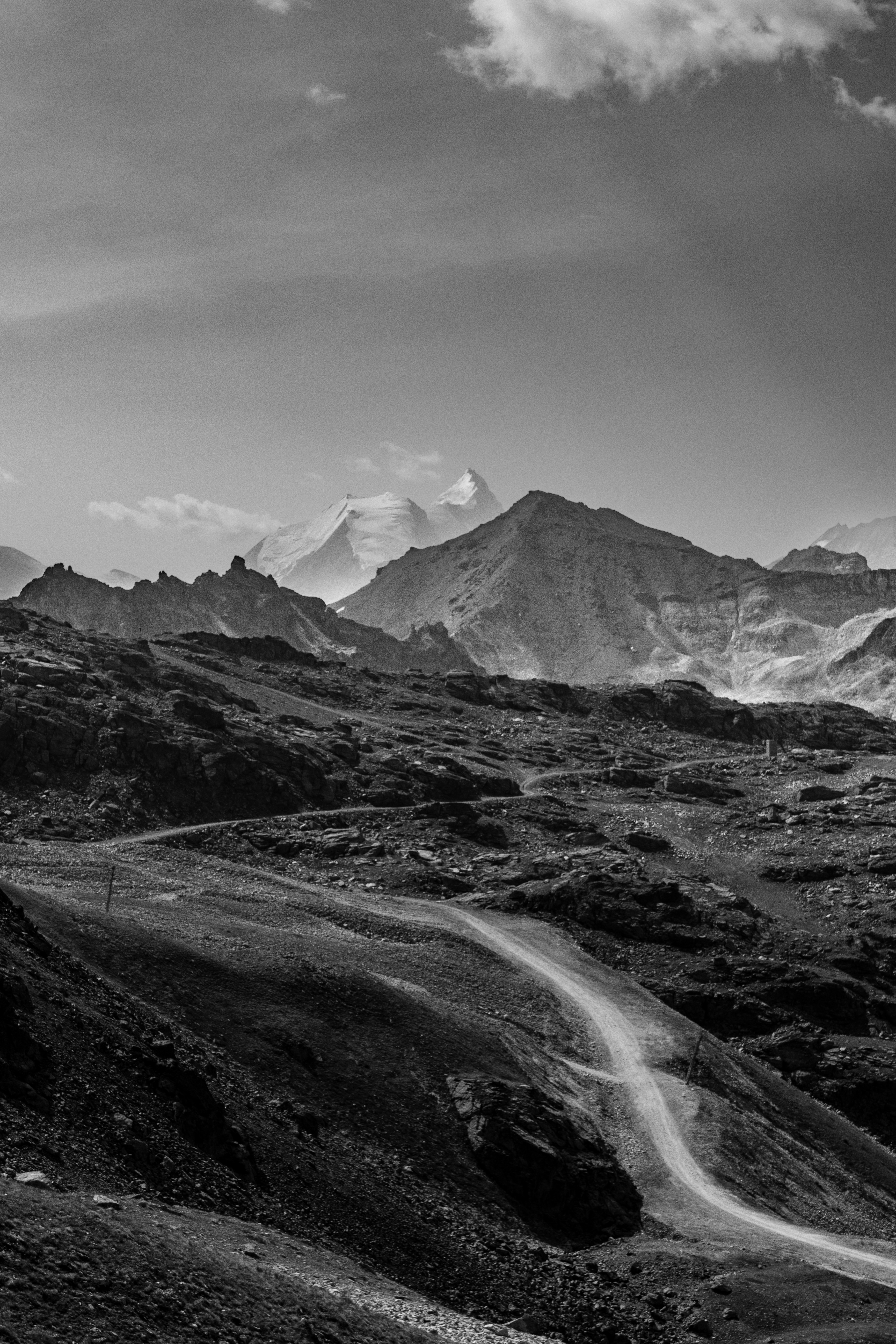 A grayscale landscape of rugged mountain ridges with a winding dirt path leading toward distant snow-cloaked peaks.