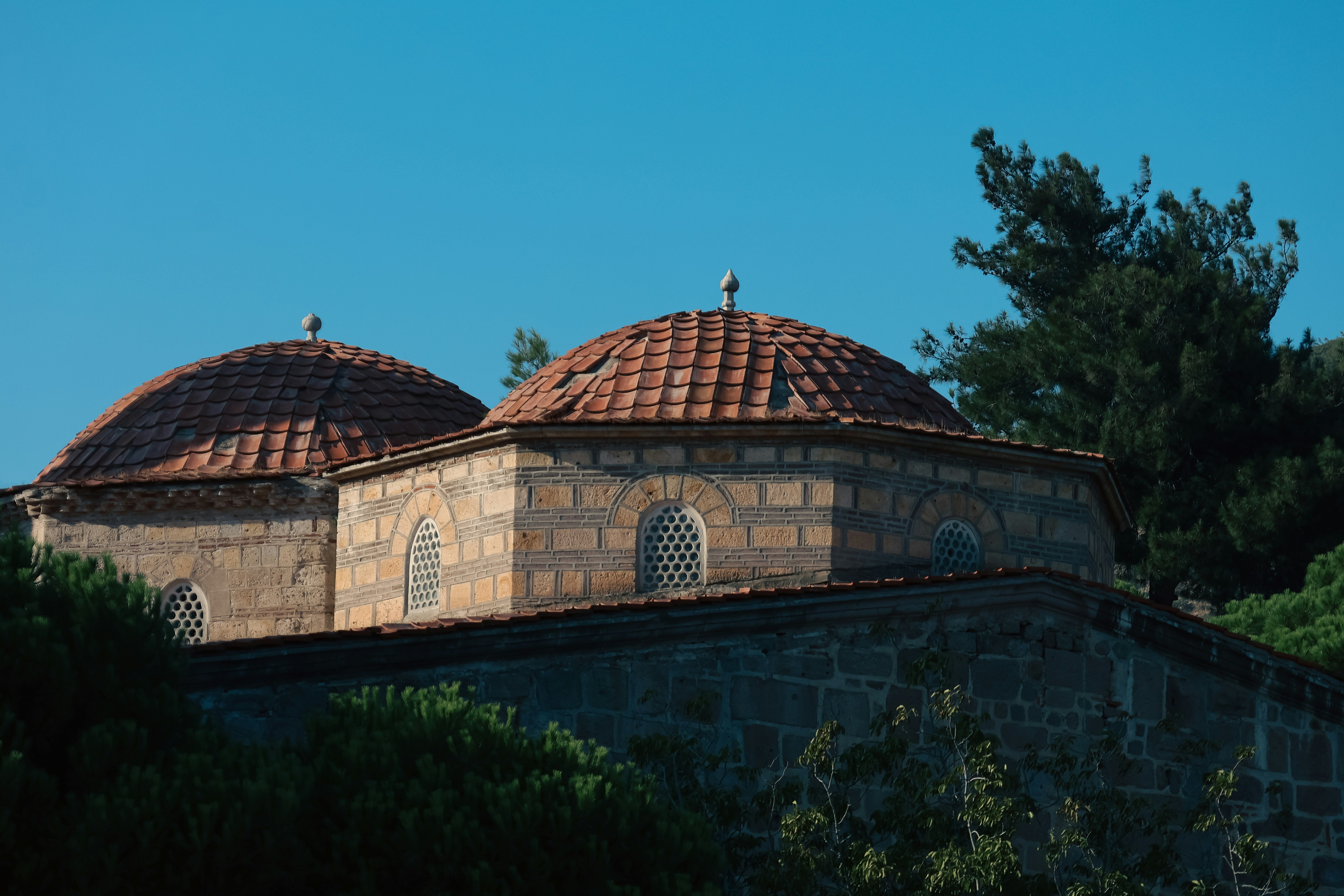 A stone building with red-tiled domes sits atop a low wall, framed by trees and a bright blue sky.