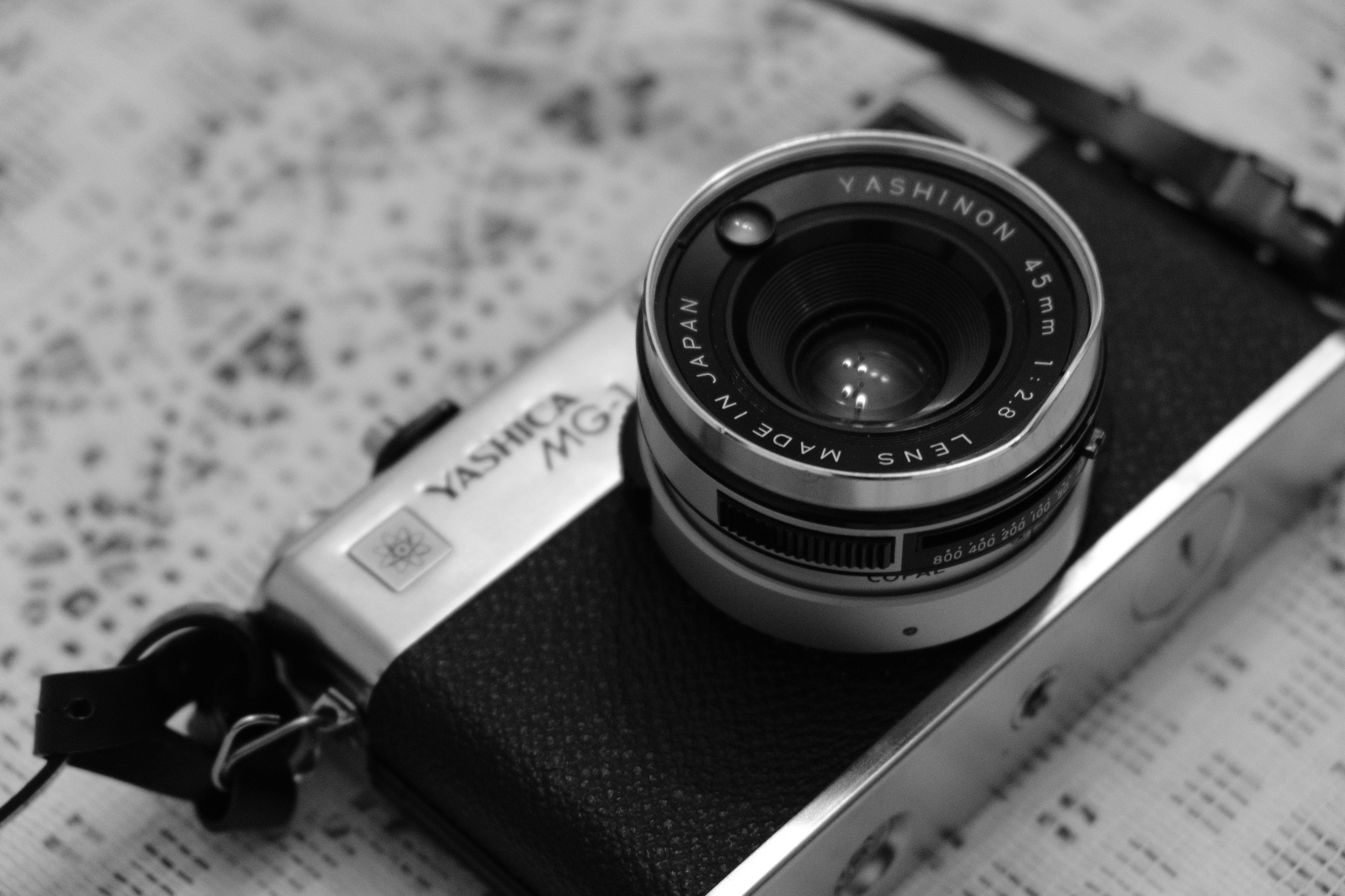 Black-and-white shot of a Yashinon 45mm lens mounted on a retro camera resting on a patterned surface, highlighting vintage hardware and tactile textures.