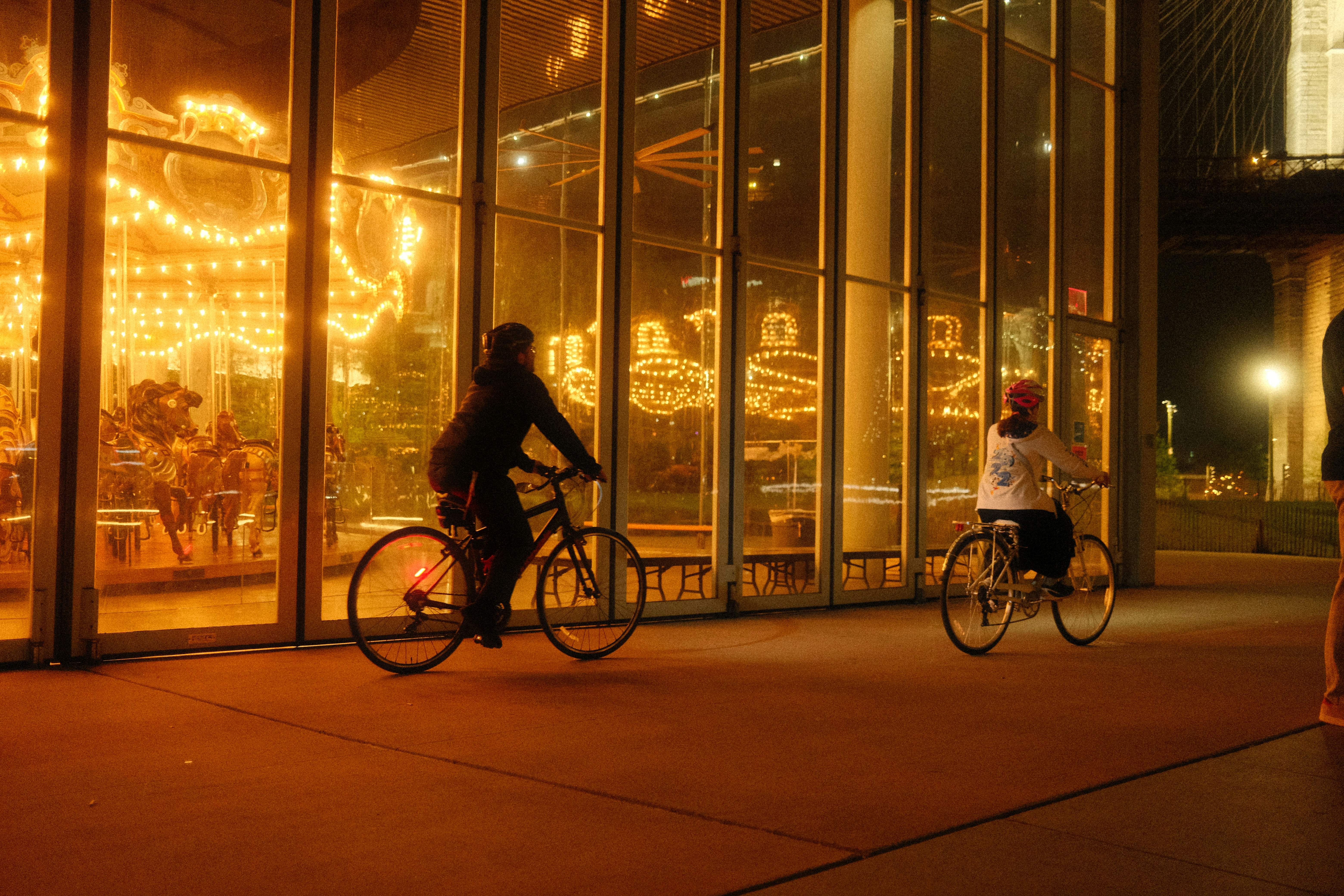 Two people cycling past a brightly lit carousel at night