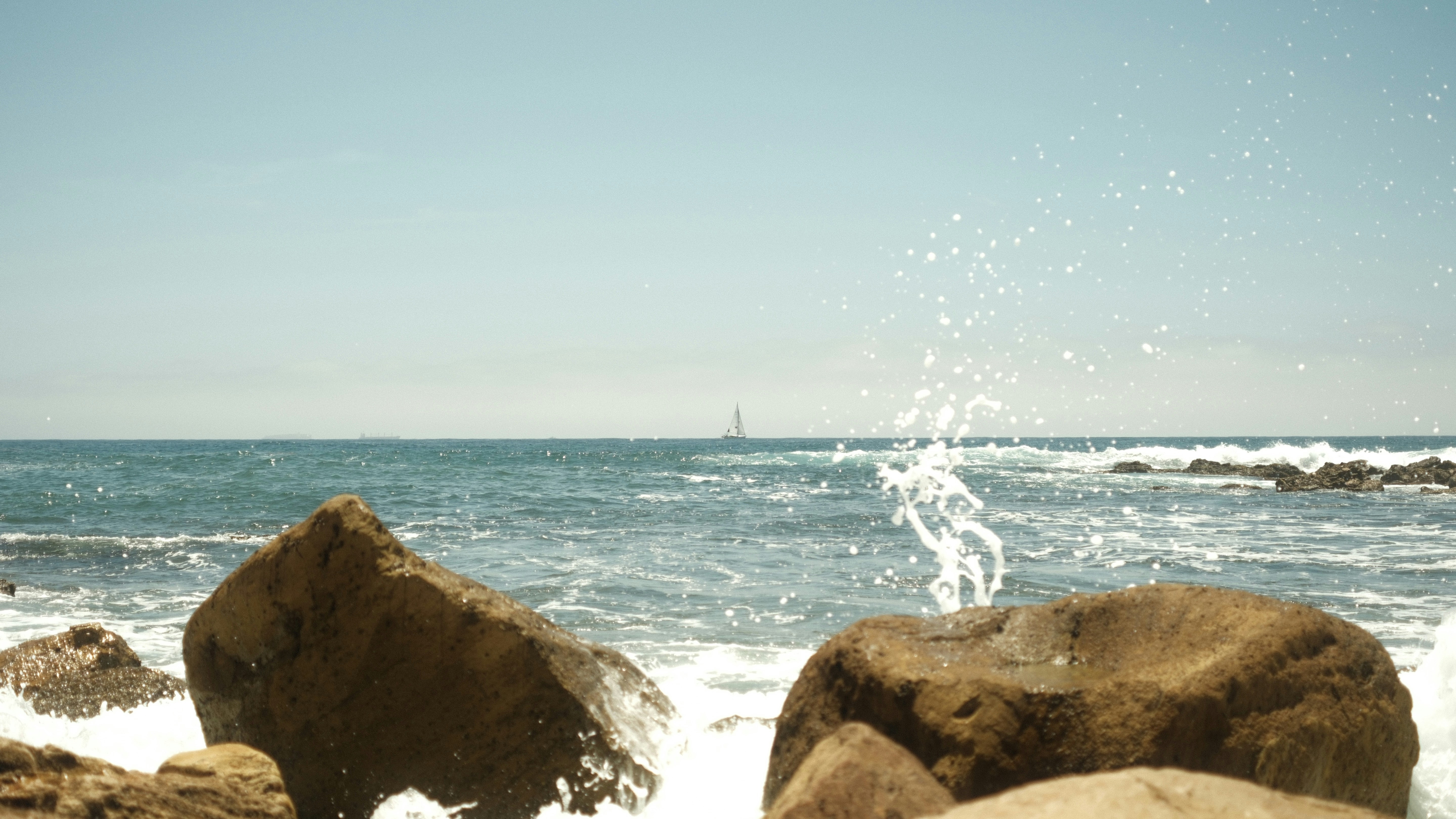 Golden-brown rocks frame a turquoise sea with white spray arching between them; a distant sailboat on the horizon anchors the tranquil coastal scene.