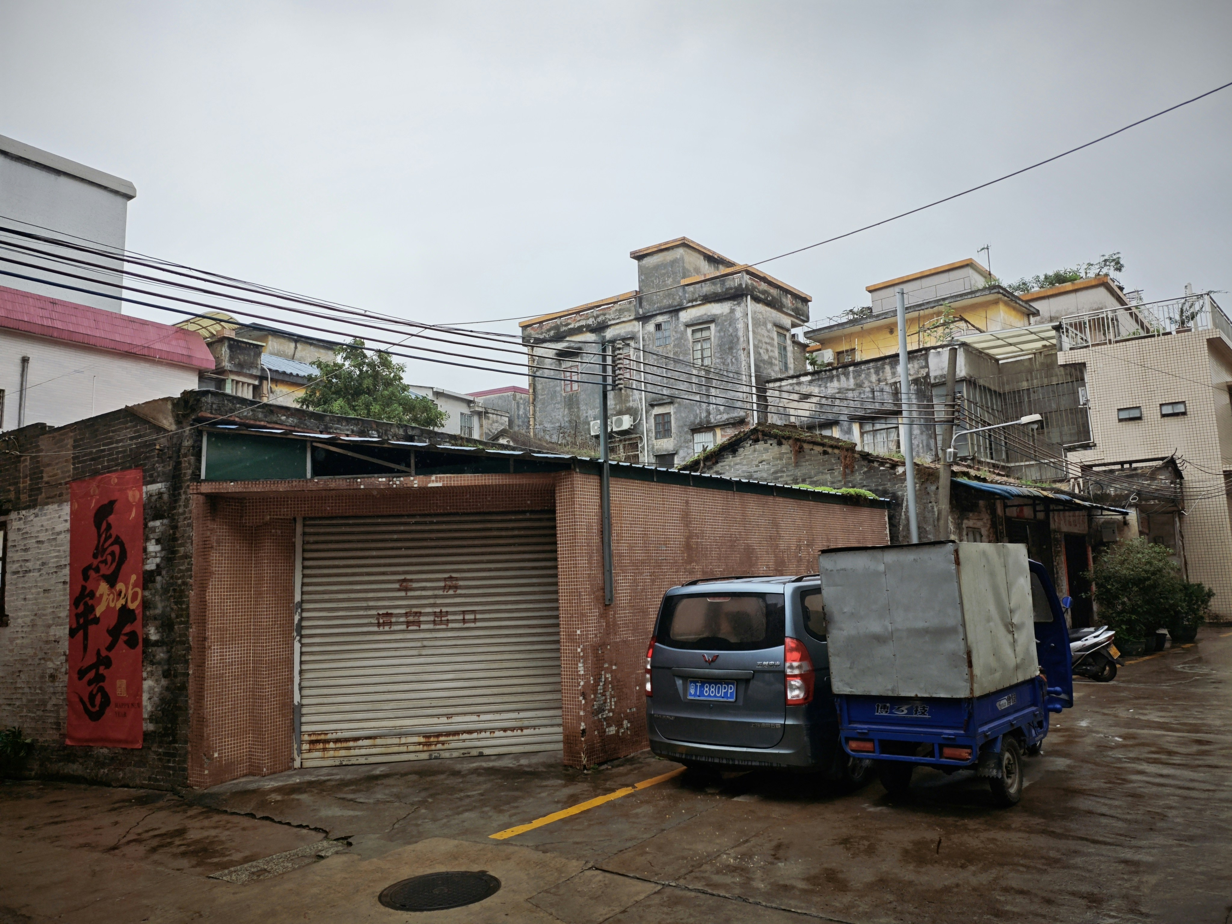 A weathered urban backstreet scene featuring a shuttered storefront, brick walls, and two parked vehicles under an overcast sky.