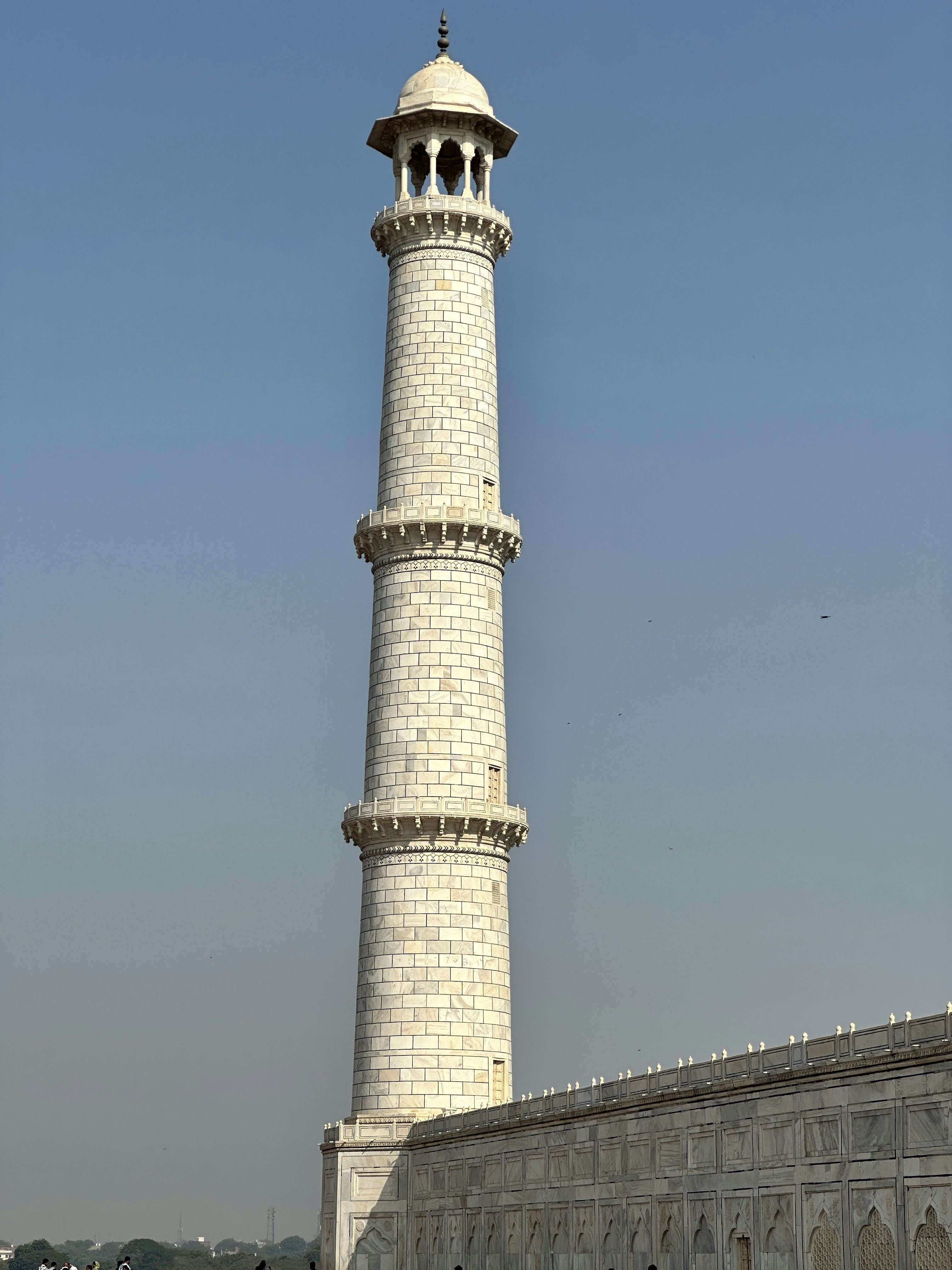 A tall white marble minaret rises beside a long stone platform under a blue sky, emphasizing monumental architecture and linear perspective.