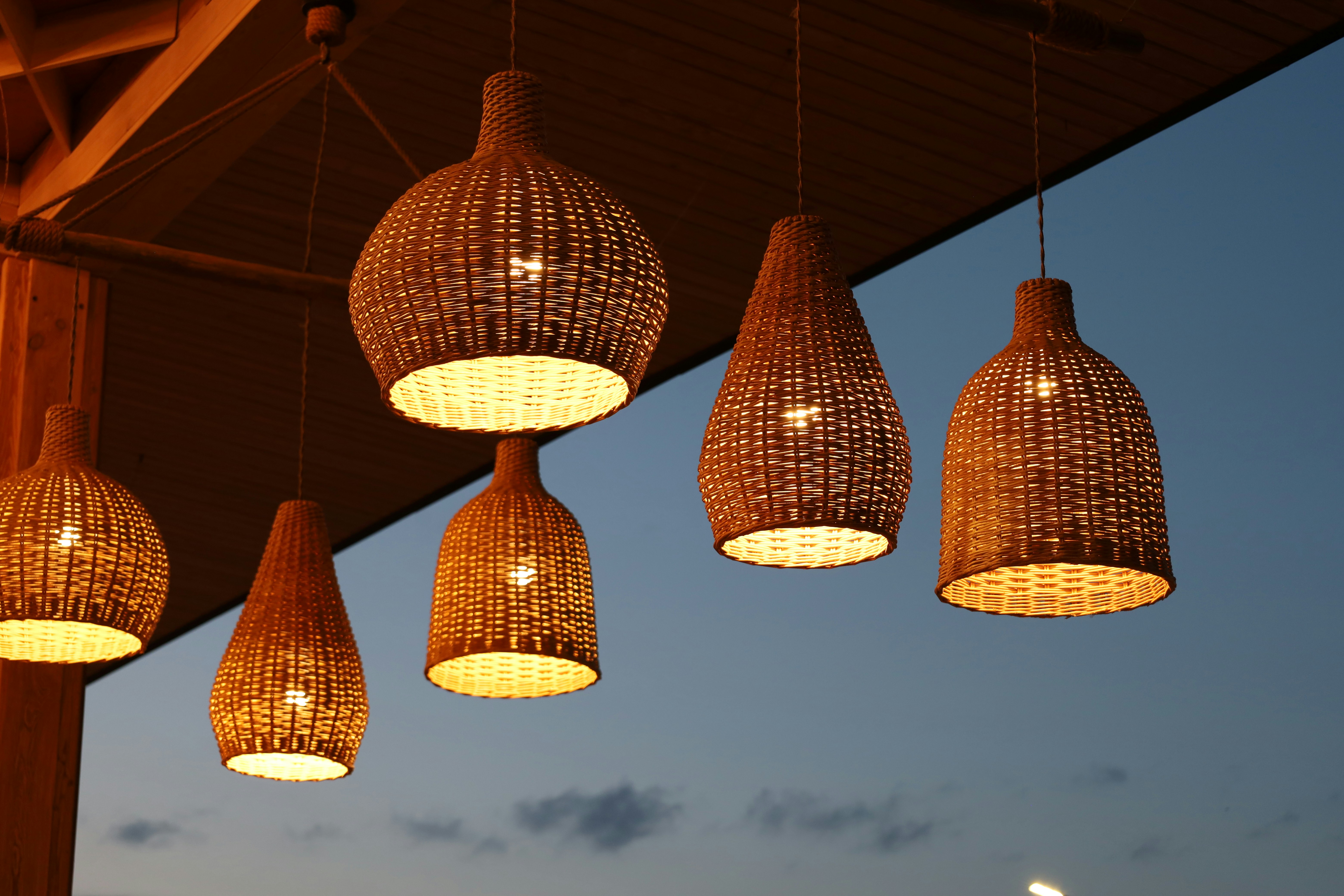 Warm woven lanterns hang from a covered patio against a dusky blue sky, their glow creating a cozy, inviting atmosphere. This is a photography capture of architectural lighting and ambient outdoor decor.
