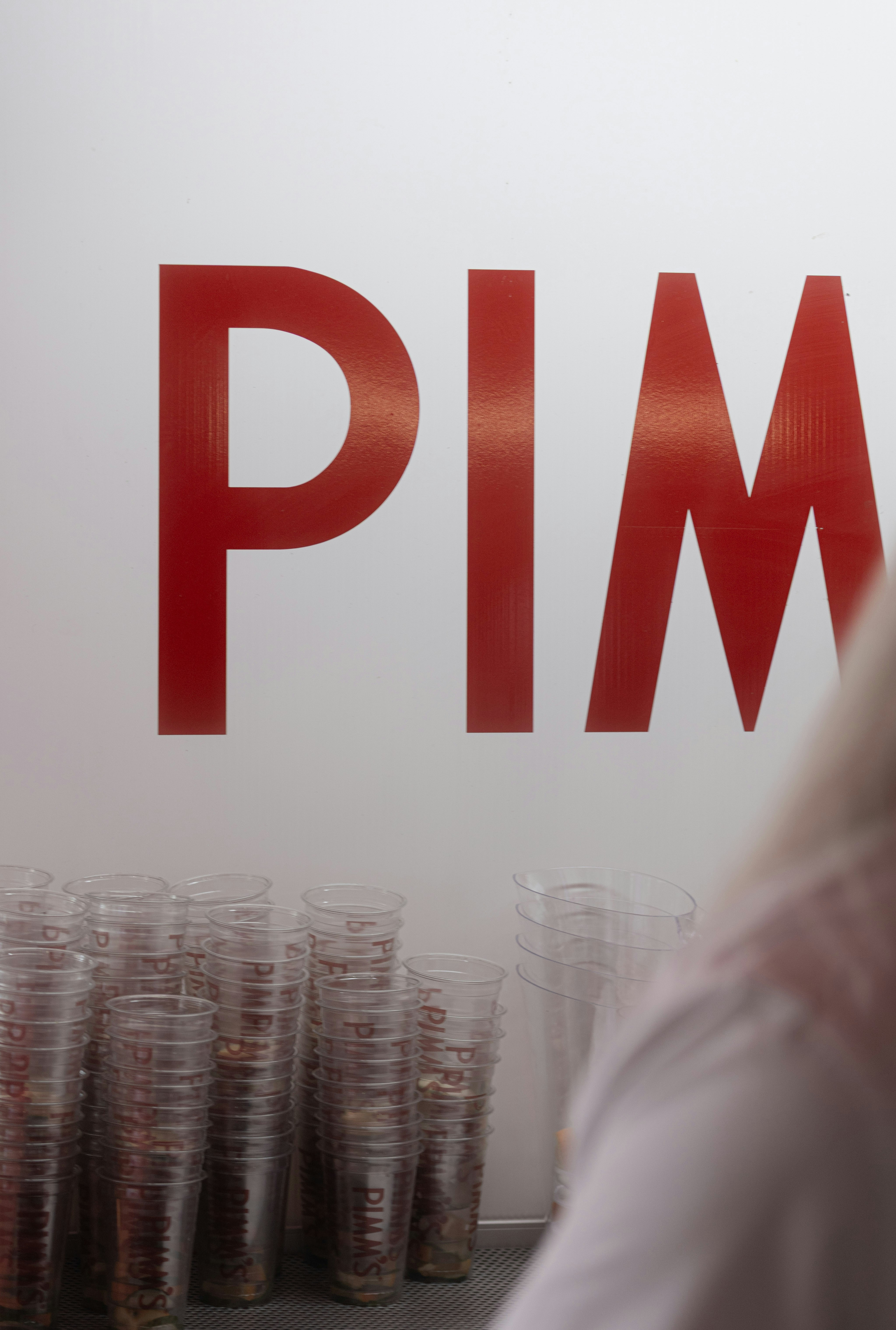 Stacked clear cups filled with beverage beneath a bold red PIM sign in a bright wall setup.