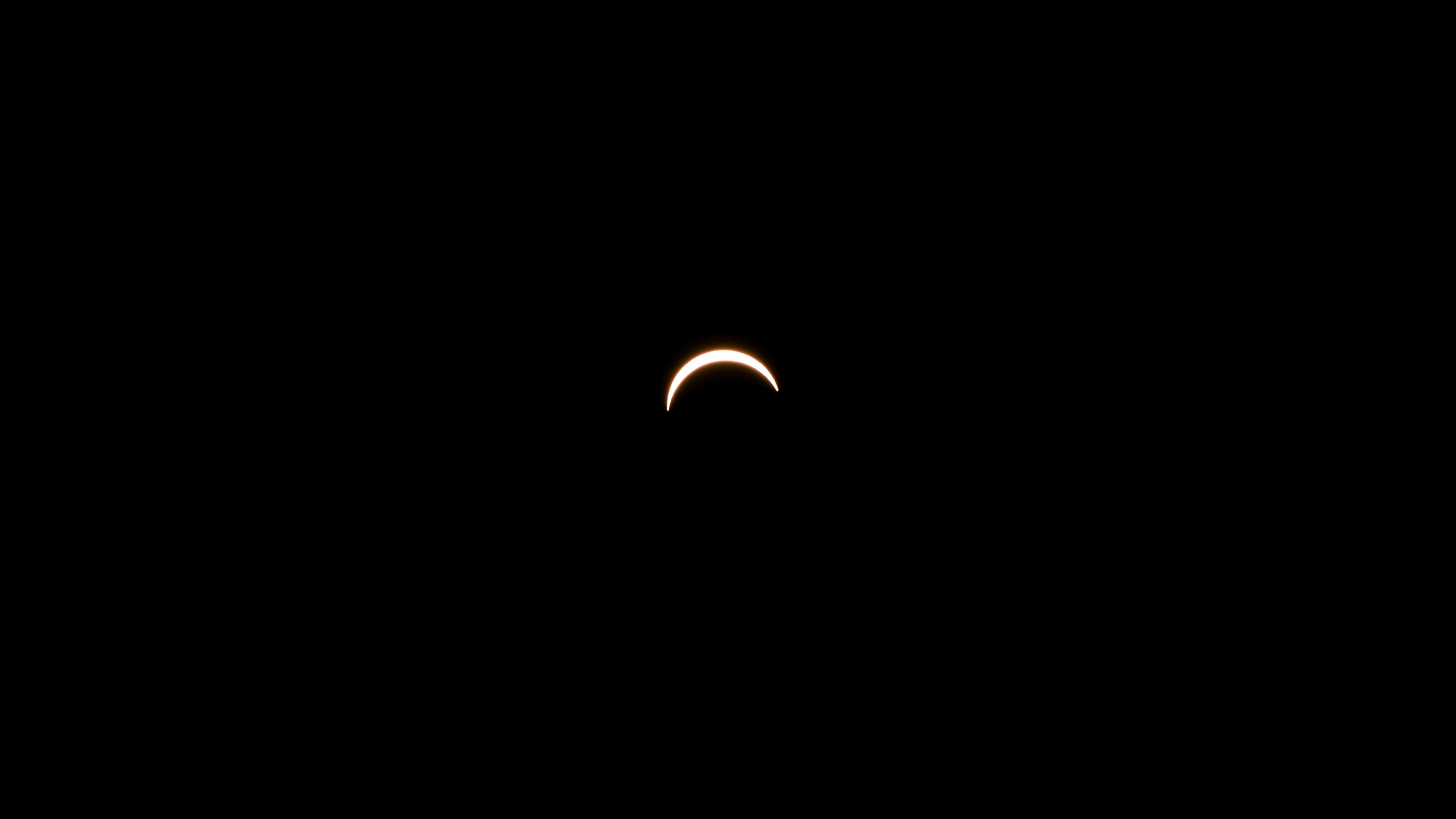 Crescent solar eclipse visible against a dark sky, showing the thin illuminated crescent formed by the sun peeking around the moon. The scene emphasizes a solitary astronomical event with high contrast.