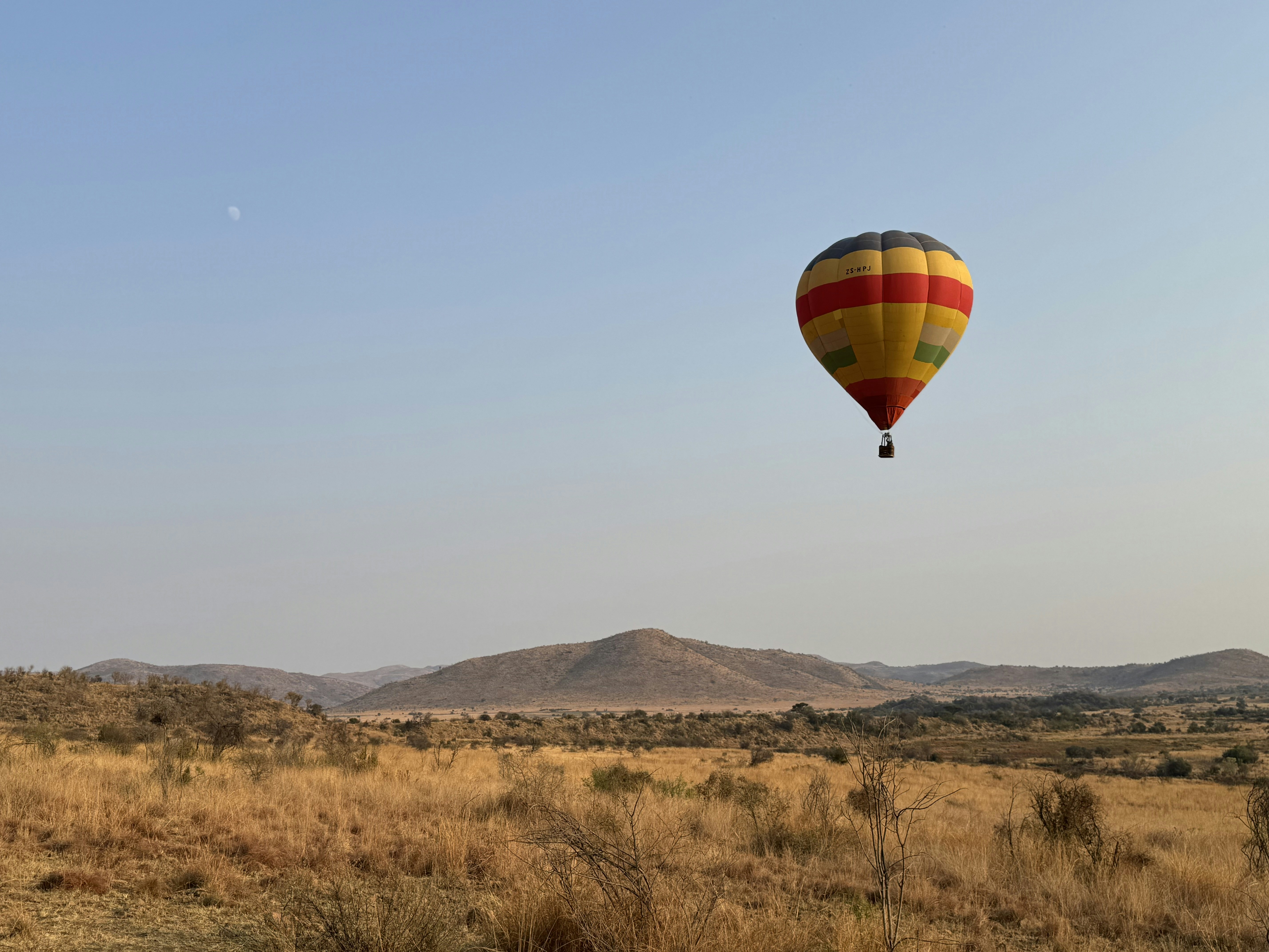 A colorful hot-air balloon drifts above a dry, open savannah with distant hills under a clear blue sky.