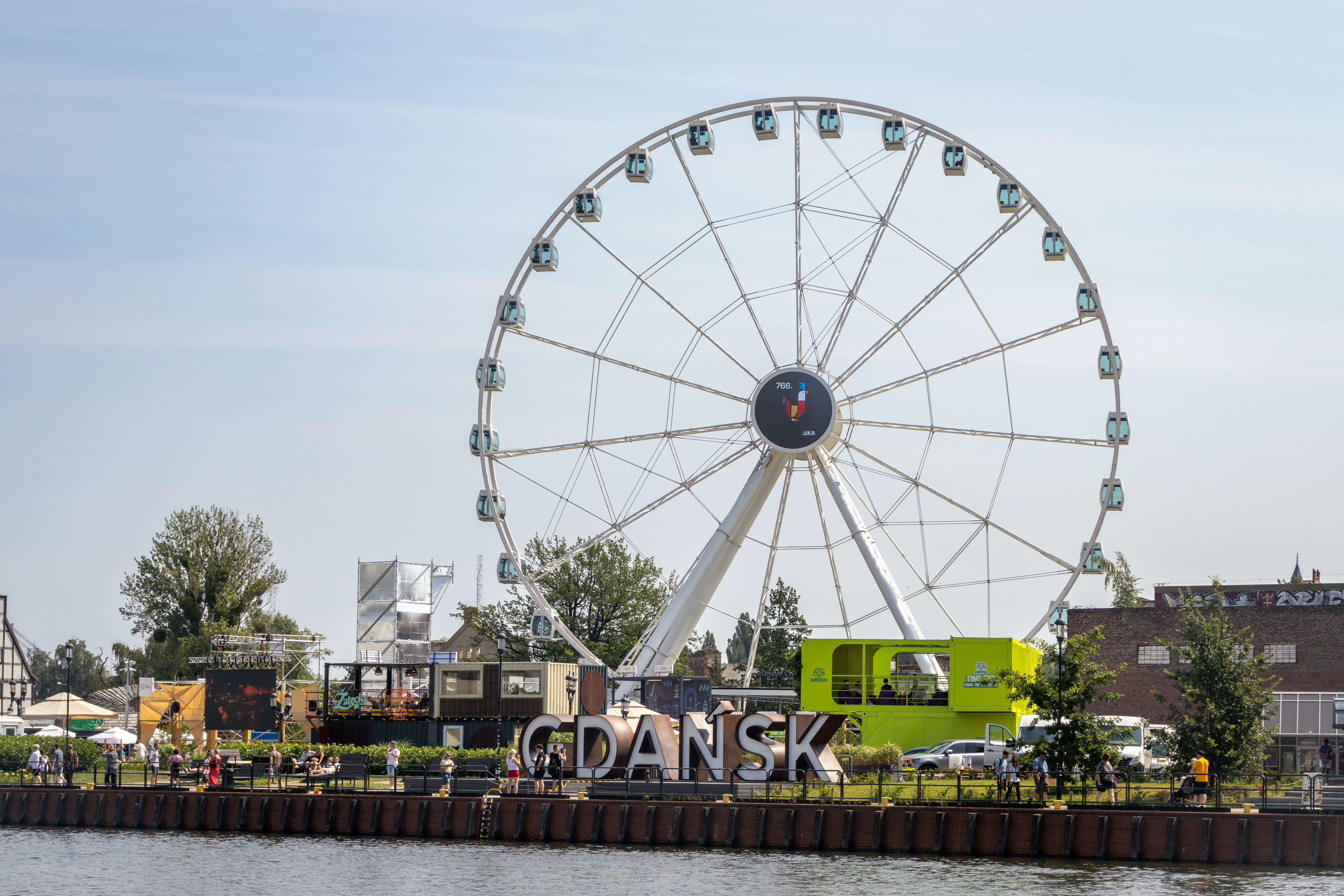 Ferris wheel dominates the skyline along a calm riverfront with a bold signage display; a lime-green vehicle and small structures line the promenade.