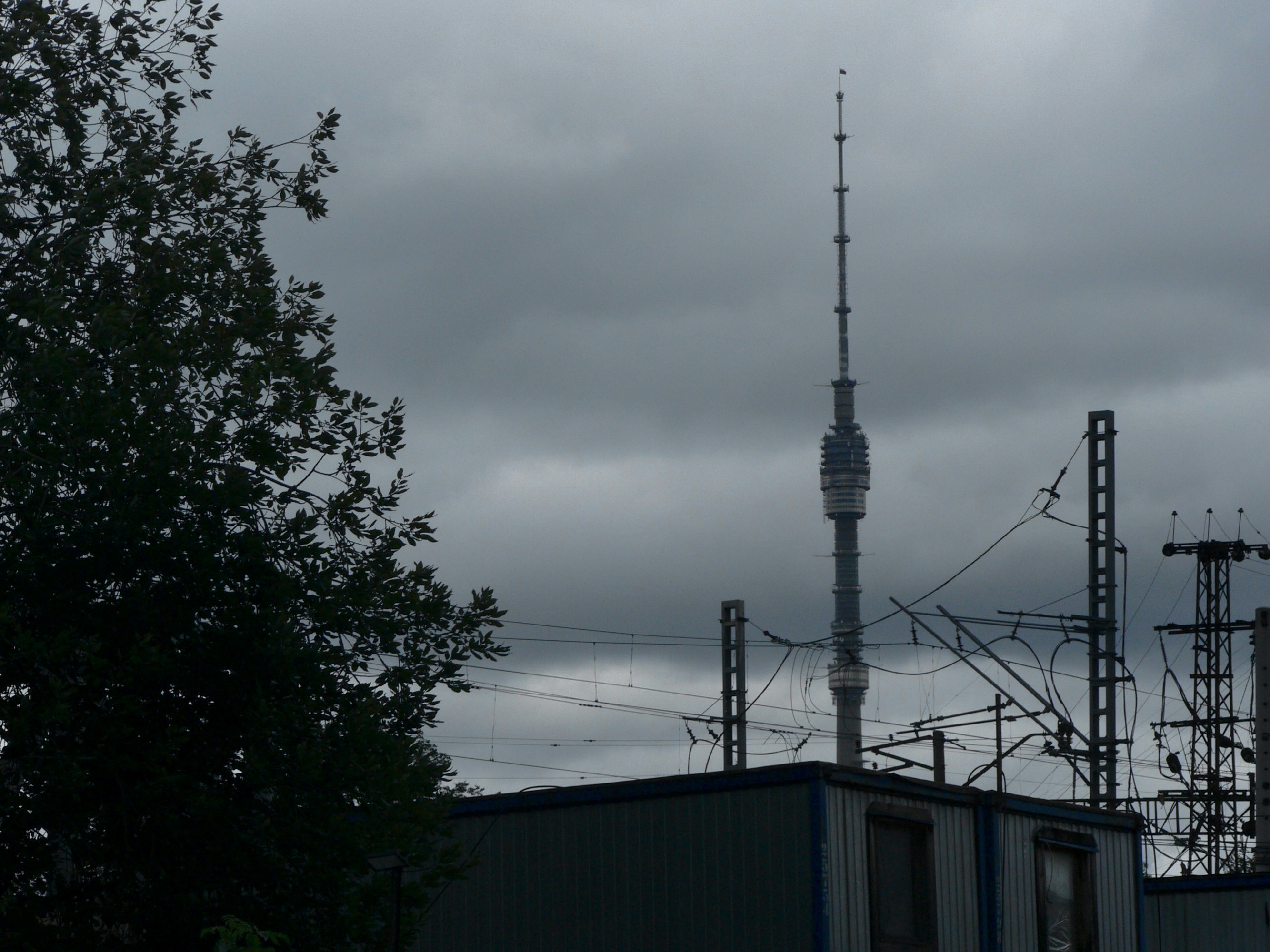 A gritty industrial urban landscape featuring the distant Ostankino TV Tower, standing tall against a gloomy grey sky. In the foreground, construction site cabins and tangled high‑voltage power lines add a raw, documentary feel to this layered view of Moscow's suburban outskirts.