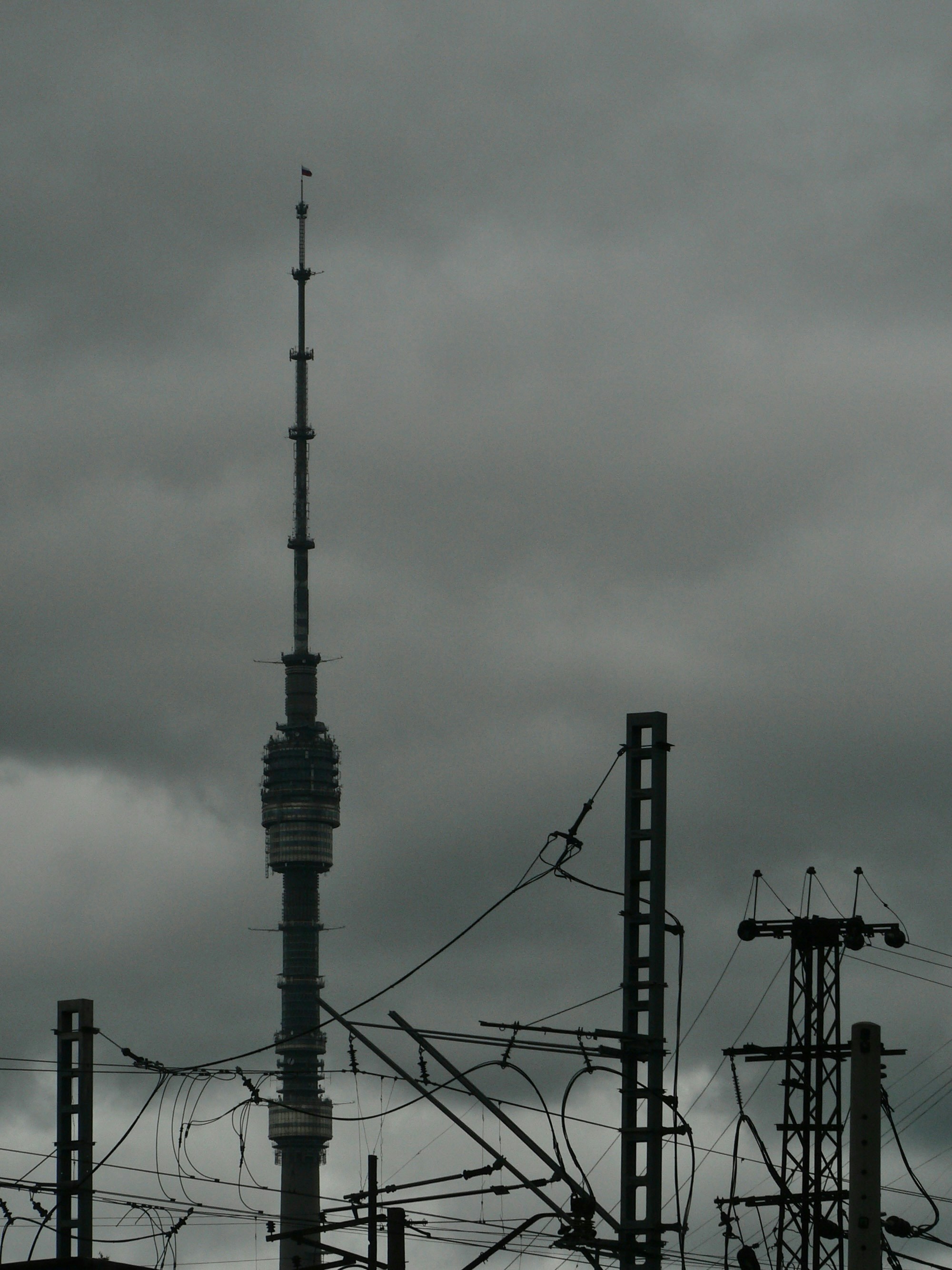 A gritty industrial urban landscape featuring the distant Ostankino TV Tower, standing tall against a gloomy grey sky. In the foreground, construction site cabins and tangled high‑voltage power lines add a raw, documentary feel to this layered view of Moscow's suburban outskirts.