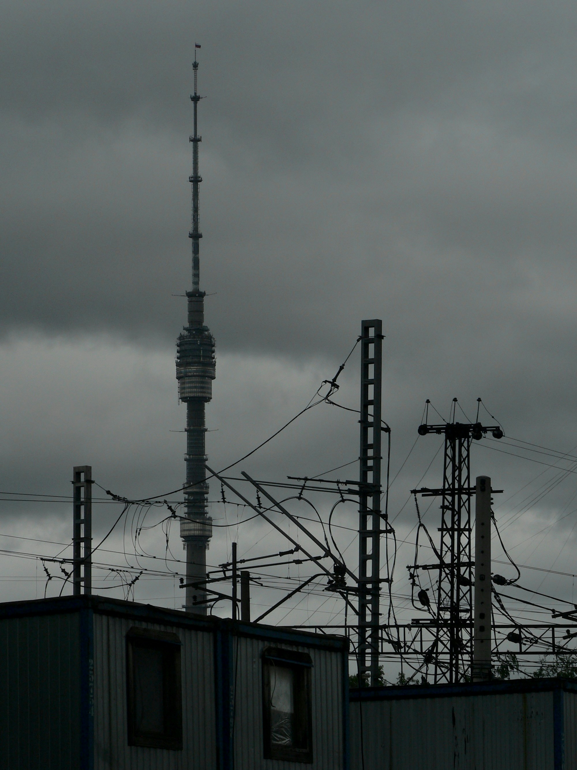 A gritty industrial urban landscape featuring the distant Ostankino TV Tower, standing tall against a gloomy grey sky. In the foreground, construction site cabins and tangled high‑voltage power lines add a raw, documentary feel to this layered view of Moscow's suburban outskirts.