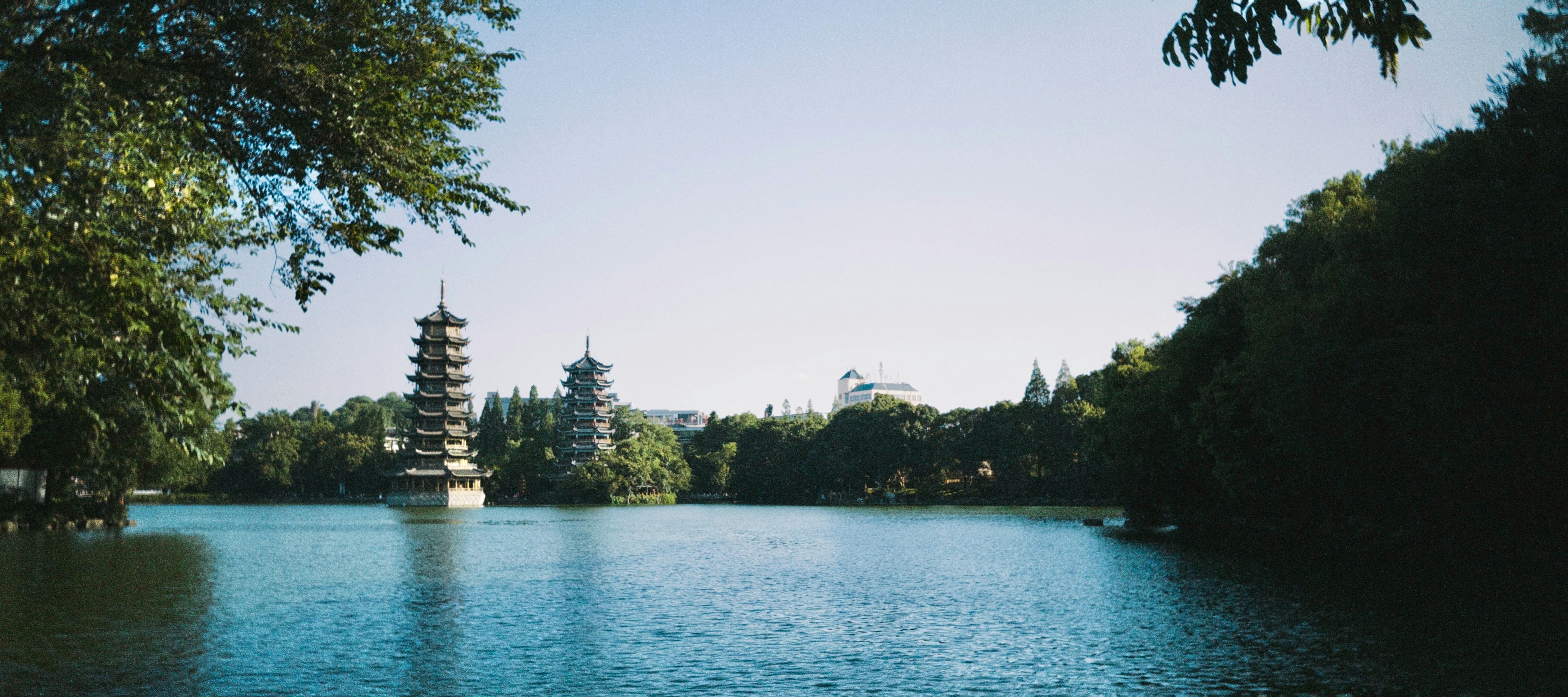 Calm lake with distant pagodas and green shoreline, captured in natural daylight. The scene emphasizes serene water and architectural elements along the horizon.