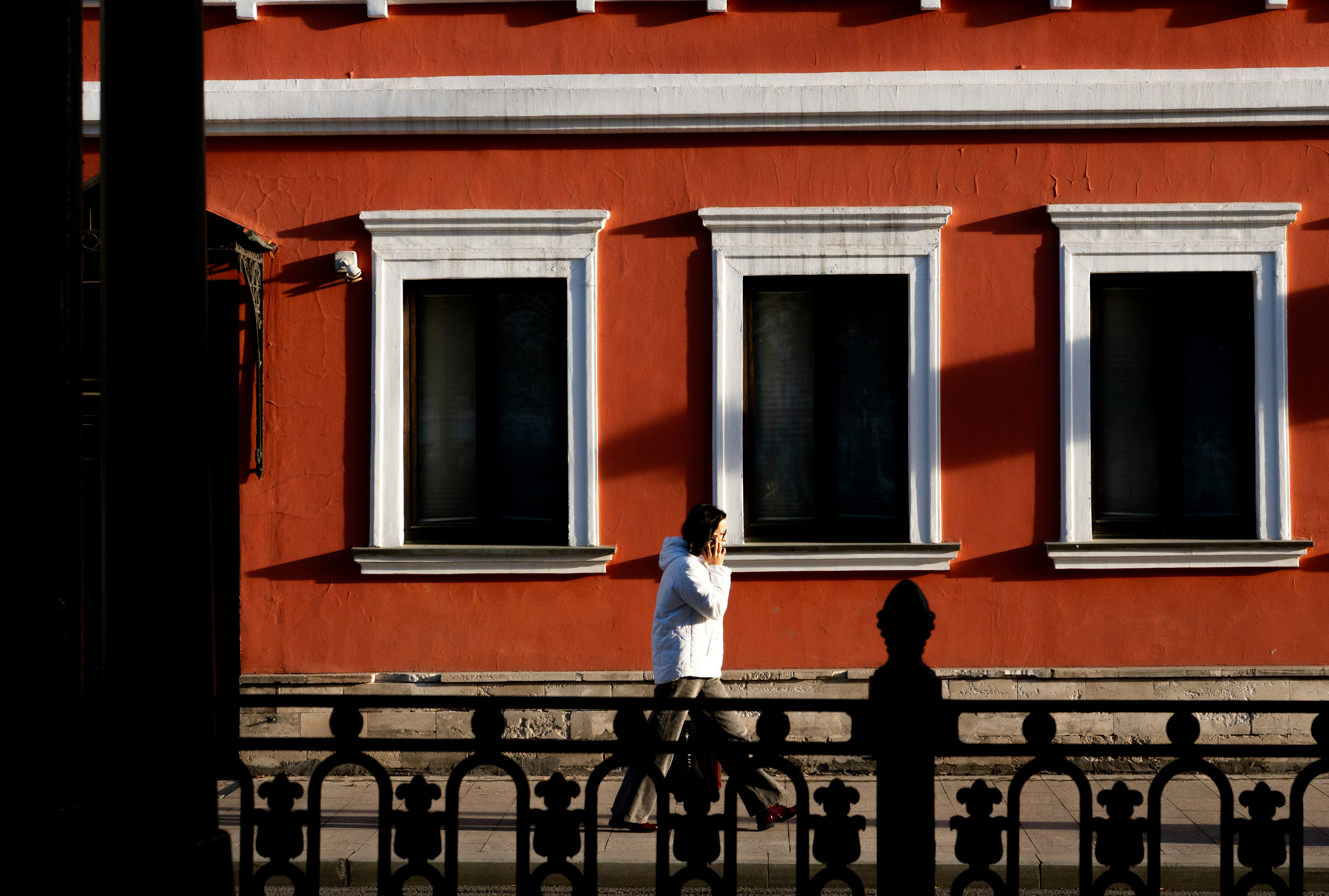 A person walking along a red-walled building casts a bold shadow on the railing, captured in bright daylight. Photograph uncertainty: a street scene with strong geometric contrast and a single passerby.