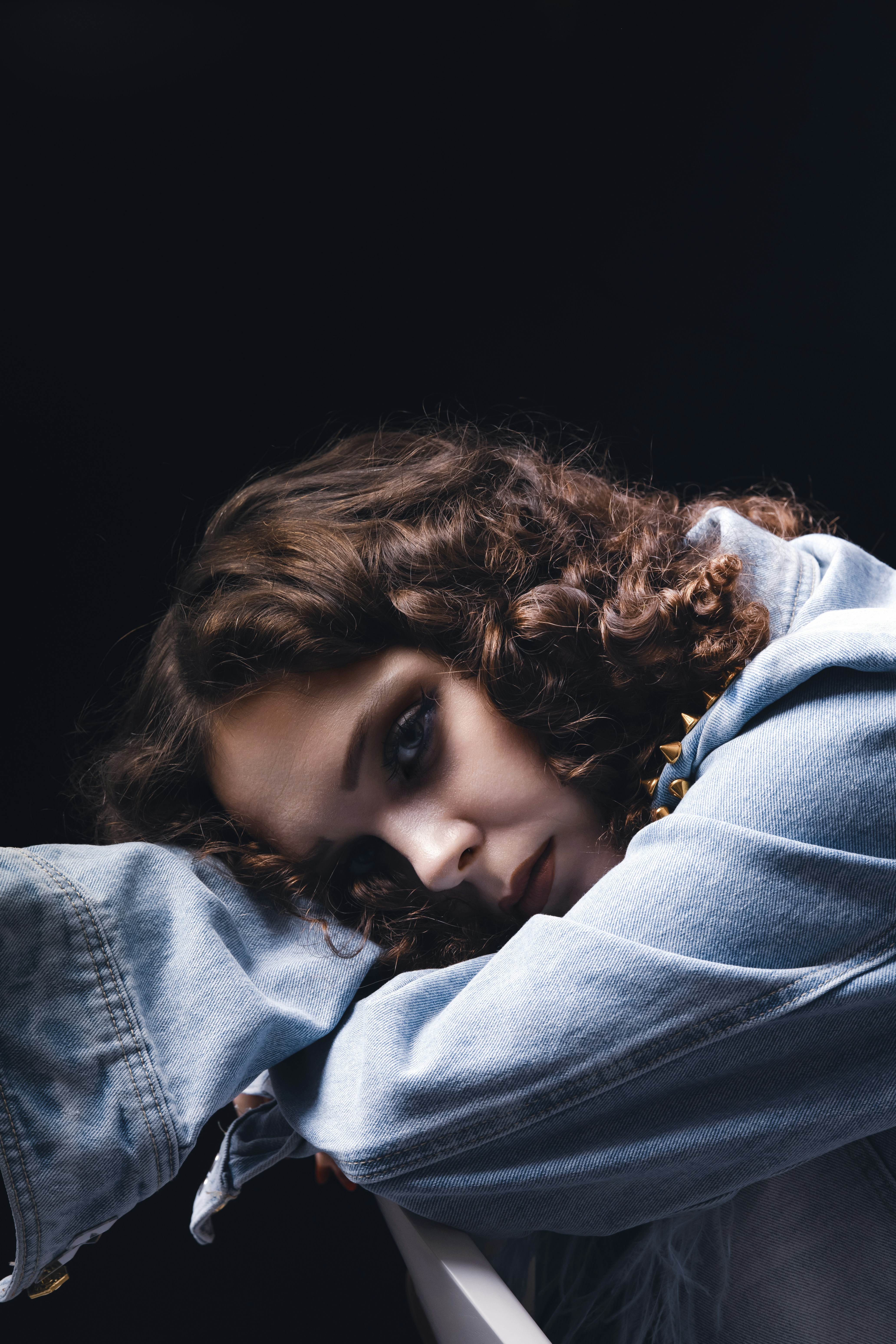 Close-up portrait of a woman with curly hair resting her head on crossed arms against a dark background; denim jacket and warm lighting define her features. This is a studio-style lifestyle photograph.