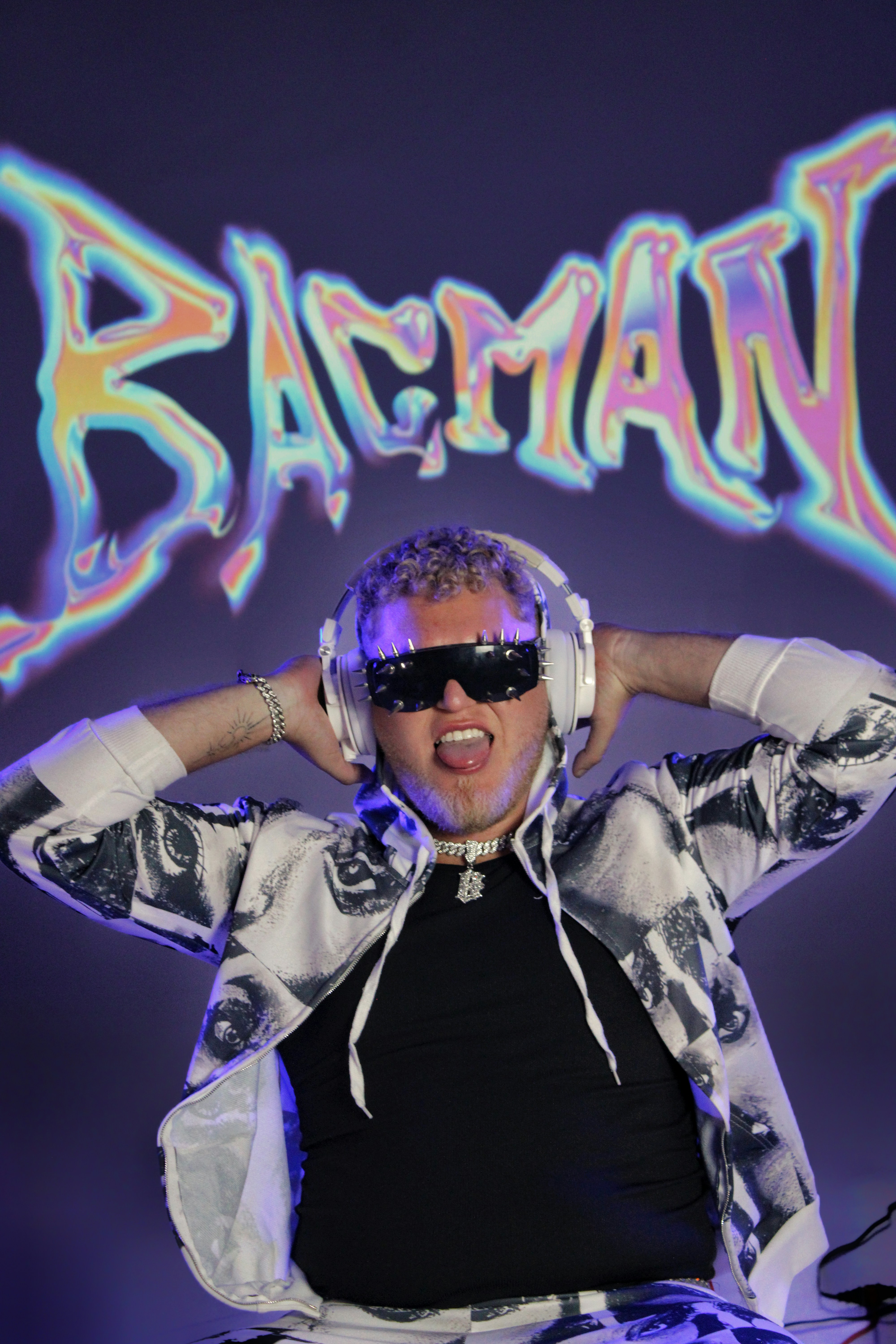 Energetic portrait of a performer wearing oversized white headphones and sunglasses, striking a relaxed, hands-behind-head pose in front of a vivid neon graffiti background. This is a high-contrast studio scene with a graphic, possibly promotional vibe.