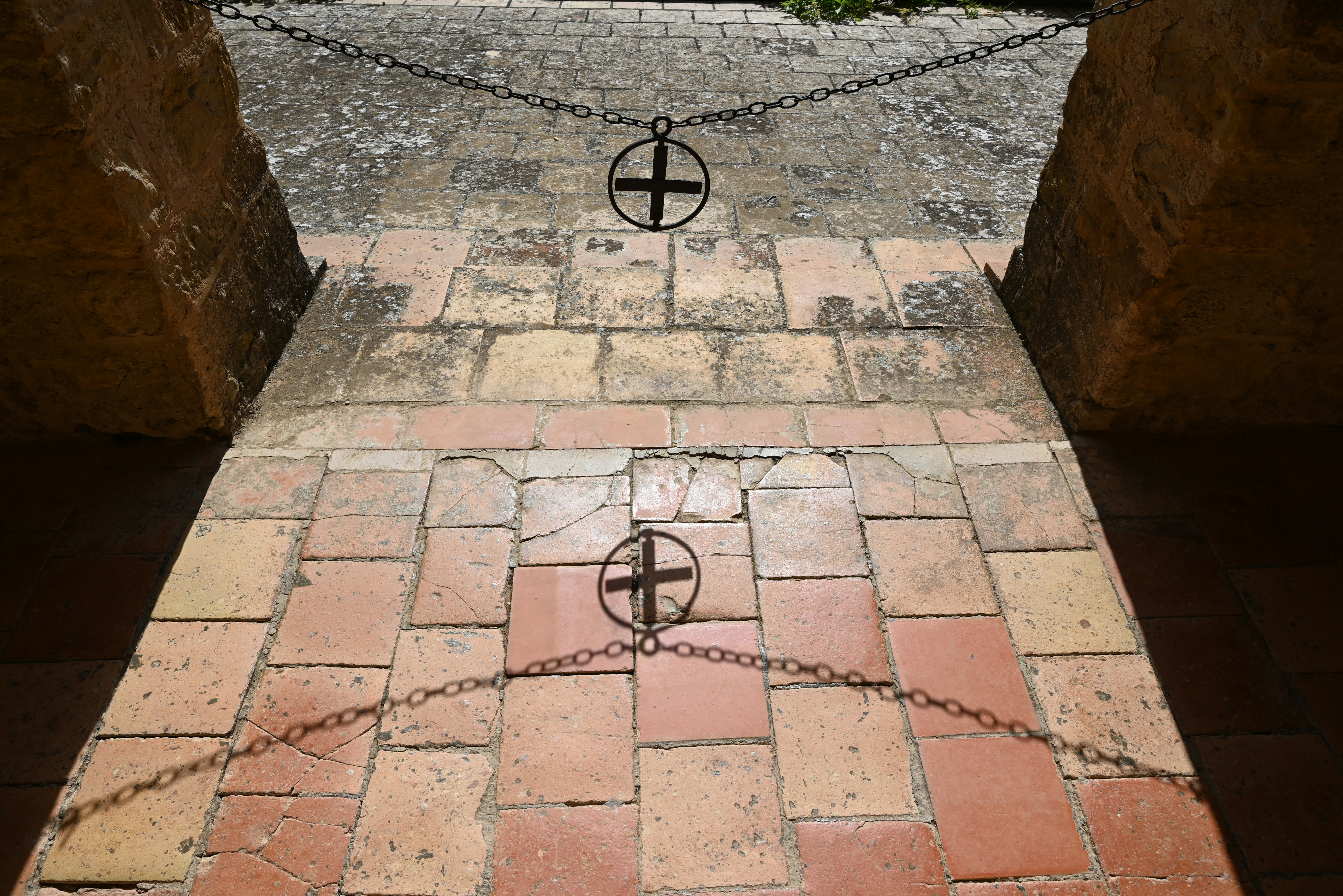 Sunlit paving leads to a chained doorway framed by stone arches, with circular metal ornaments creating subtle reflections on the brick floor.