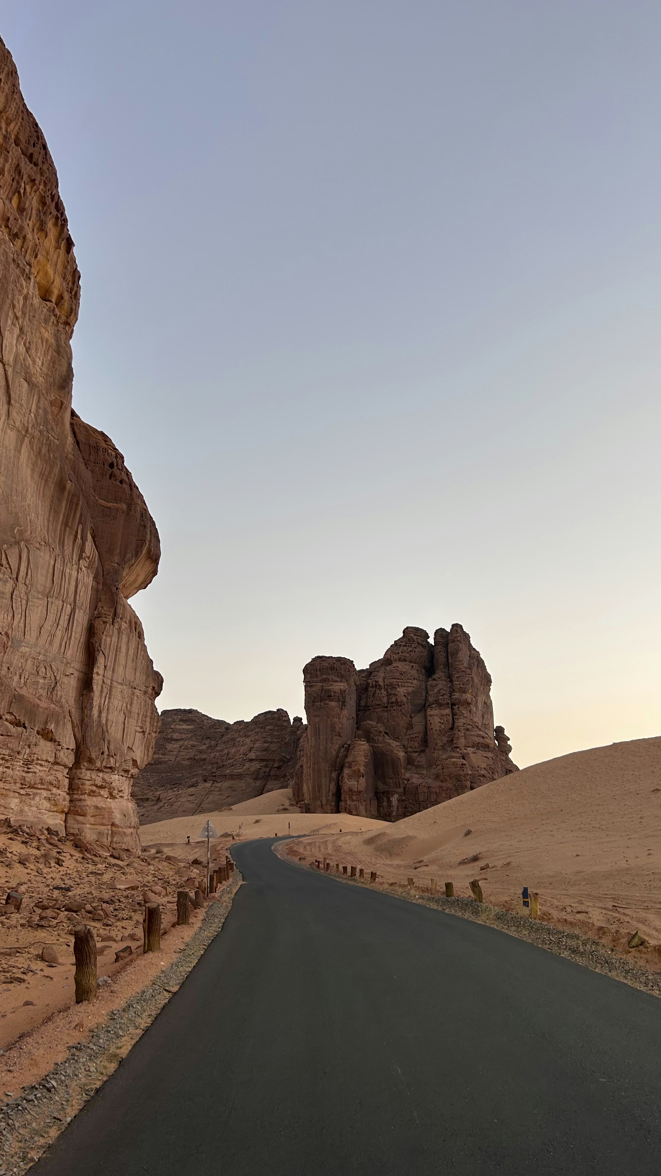 A winding paved road threads between towering rock formations in a desert canyon, with warm dunes on the right and a distant rock outcrop under a clear sky.