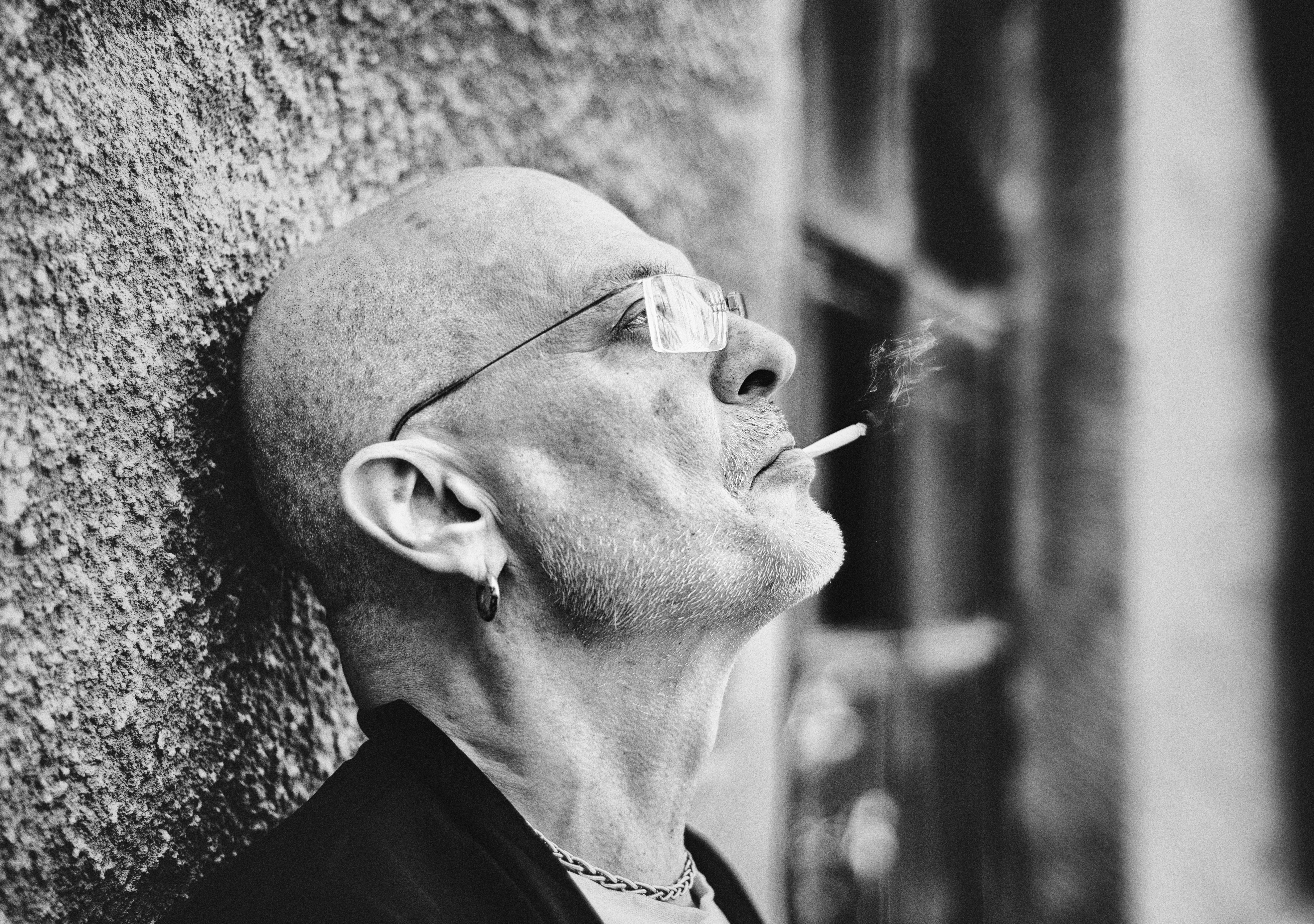 Bald man with cigarette and sunglasses rests against a textured wall in a moody black-and-white street portrait. The shot emphasizes rugged character and urban grit.