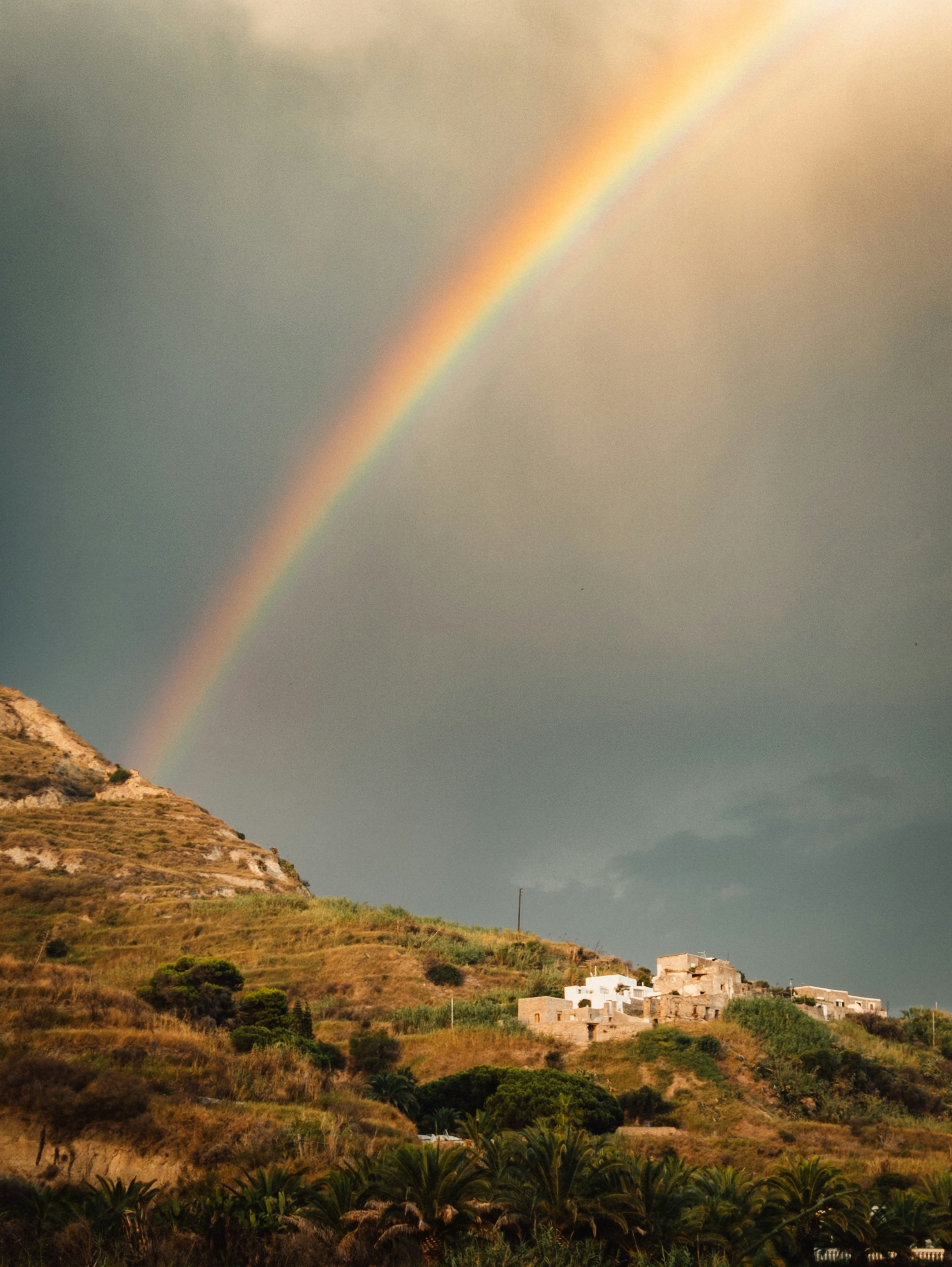 A bright rainbow arching over a hillside with stone buildings and palm trees
