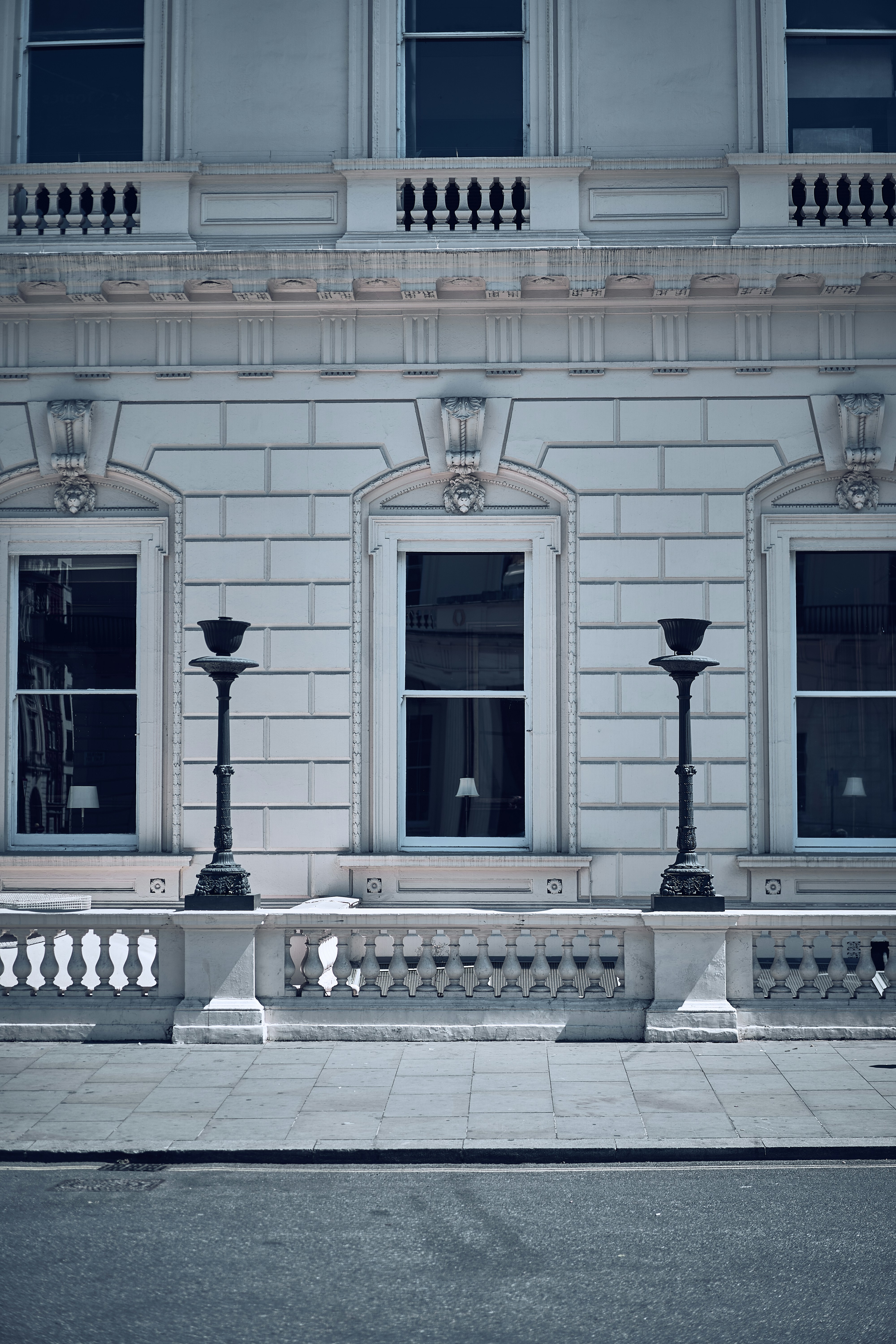 Symmetrical neoclassical facade with three windows and ornate street lamps along a balustrade; the scene emphasizes architectural details and calm street-level curb.