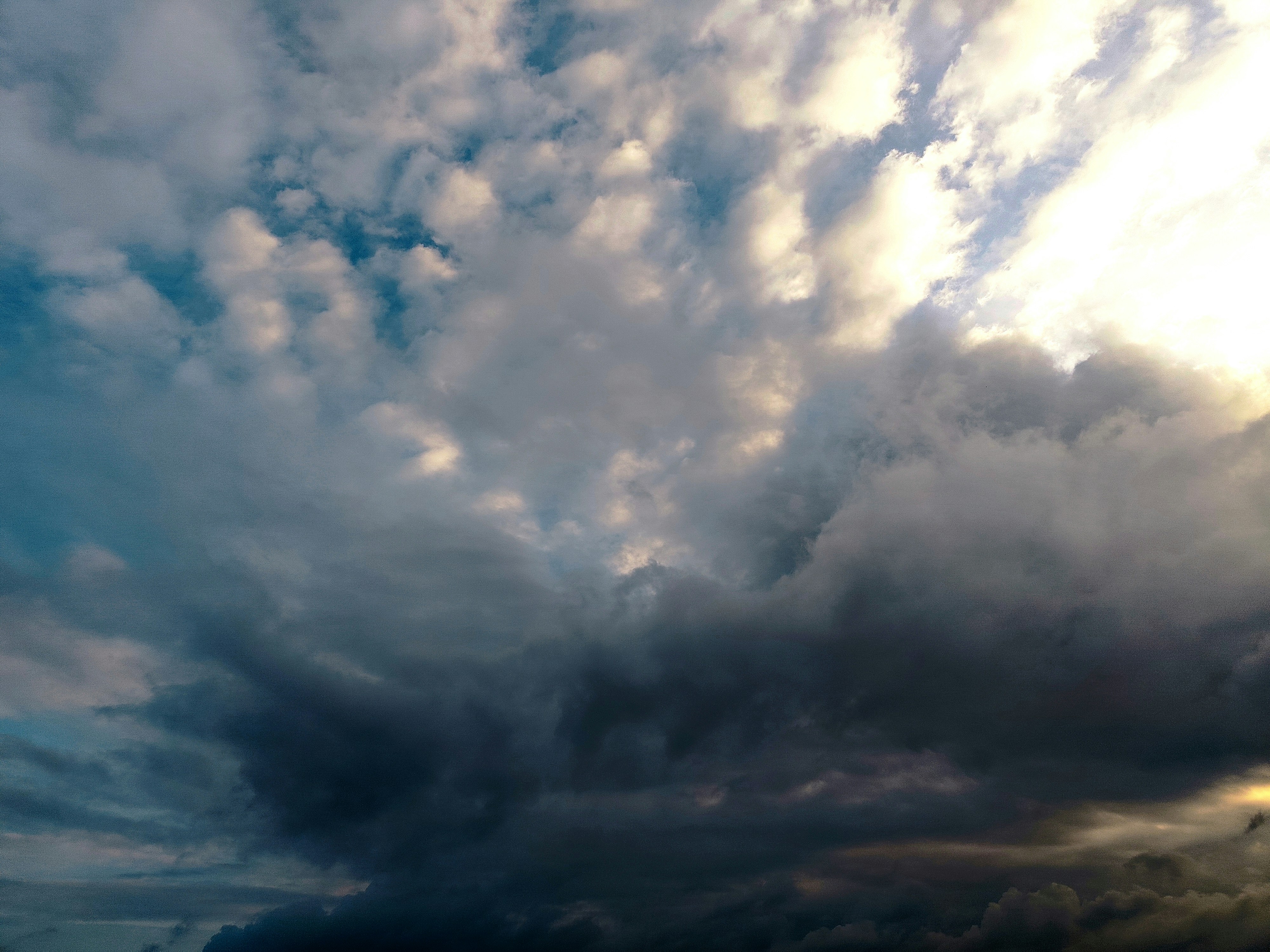 A dramatic cloudy sky featuring dark, moody storm clouds below contrasting with bright, sunlit cloud textures breaking through a soft blue sky above.