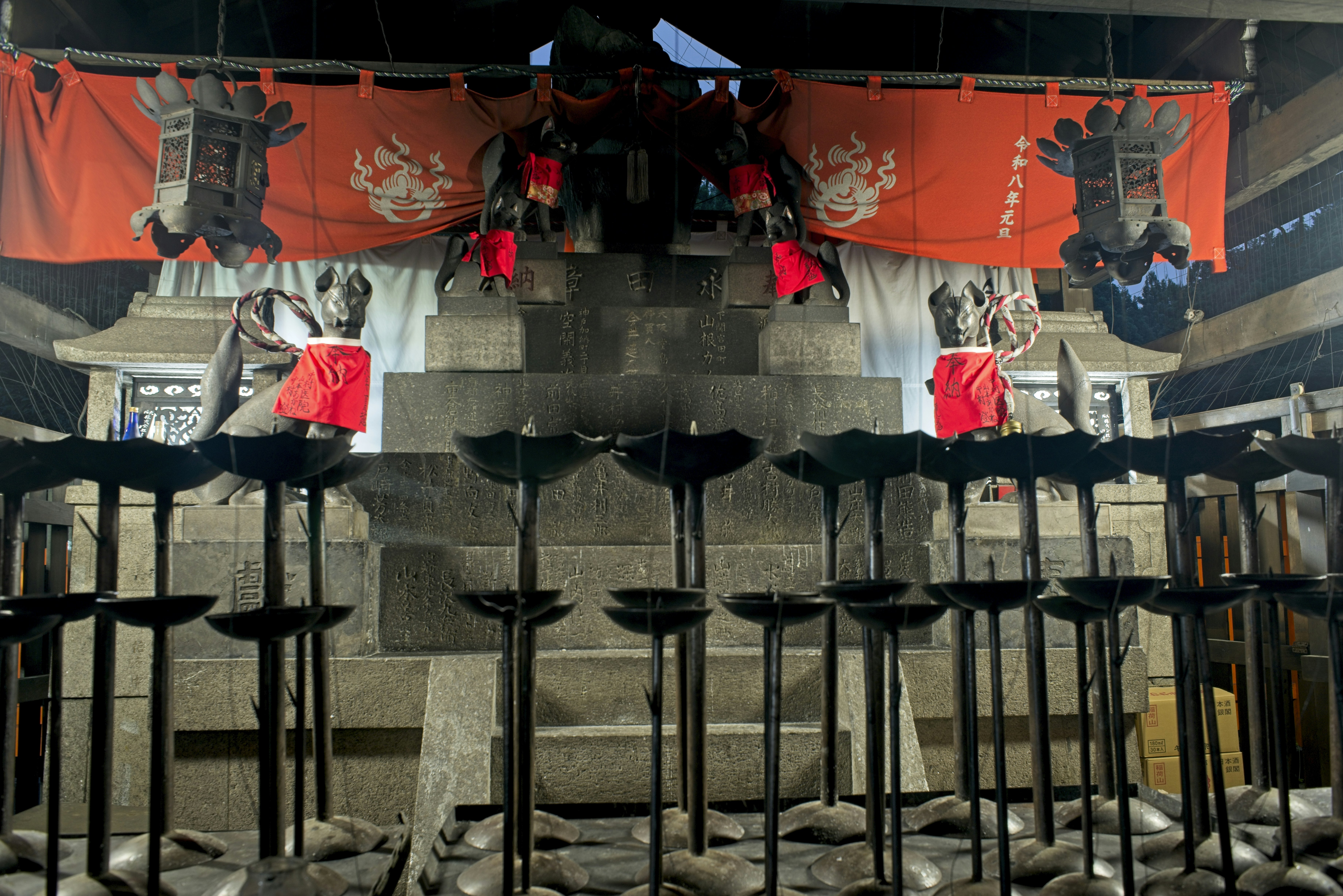 A long exposure captures four stone kitsune statues, each adorned with red ceremonial bibs, guard a densely inscribed stone altar at a subsidiary shrine along the Fushimi Inari Taisha trail, Kyoto. A vibrant red curtain bearing cloud motifs and the date "令和八年元旦" hangs above bronze lanterns, while rows of iron candle stands line the foreground. The intricate detailing and layered offerings reflect the deep devotional tradition surrounding Inari worship at this iconic Kyoto shrine.