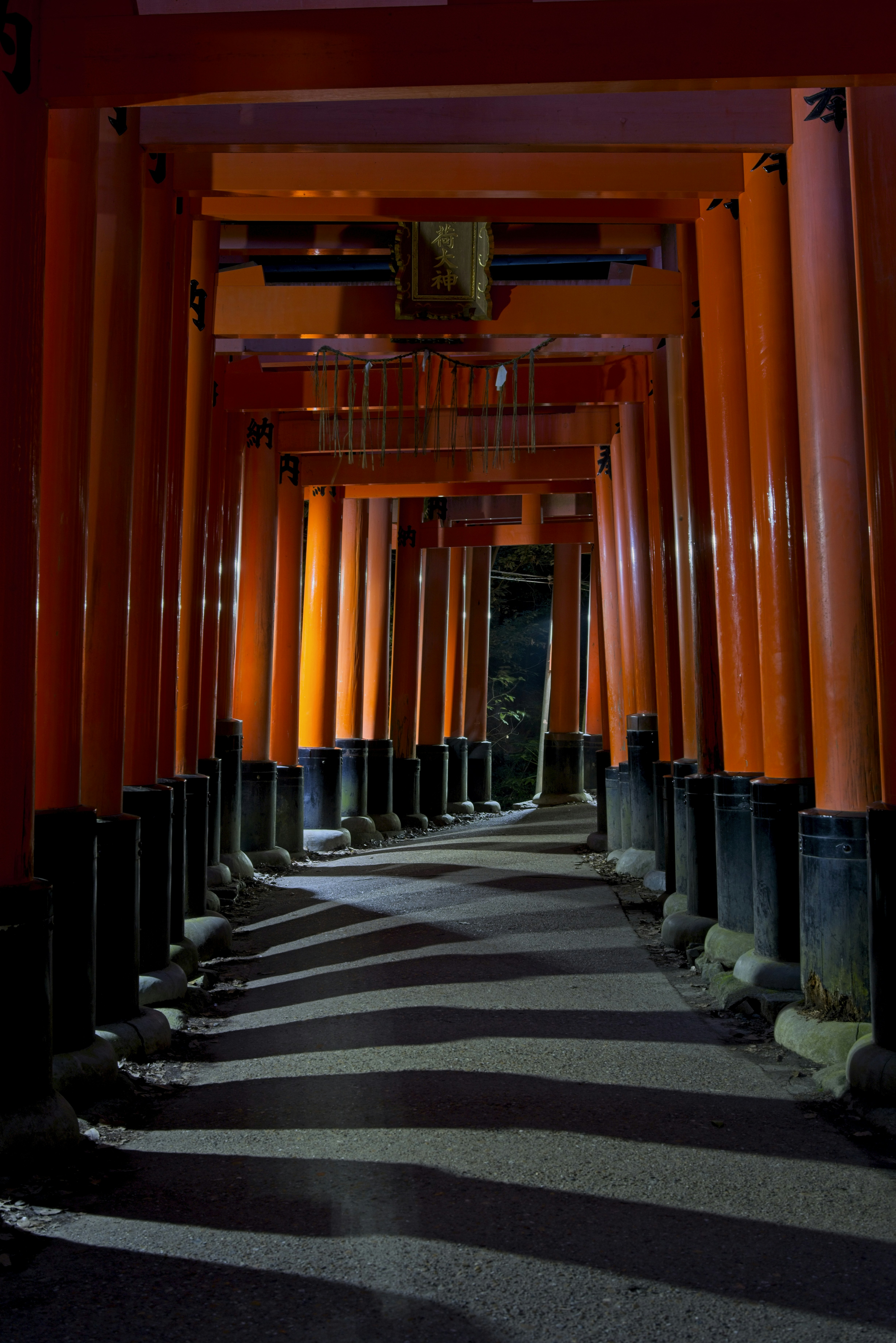 A long exposure captures an empty stretch of vibrant vermillion torii gates creates a striking tunnel-like passage at Fushimi Inari Taisha, Kyoto, illuminated softly against the darkness of a humid July evening. A sacred shimenawa rope hangs beneath a golden inscribed plaque, while striped shadows fall across the stone pathway leading into the shadowed forest beyond. The solitude and repeating structure of the gates evoke the quiet, meditative atmosphere of this iconic Kyoto shrine after nightfall.