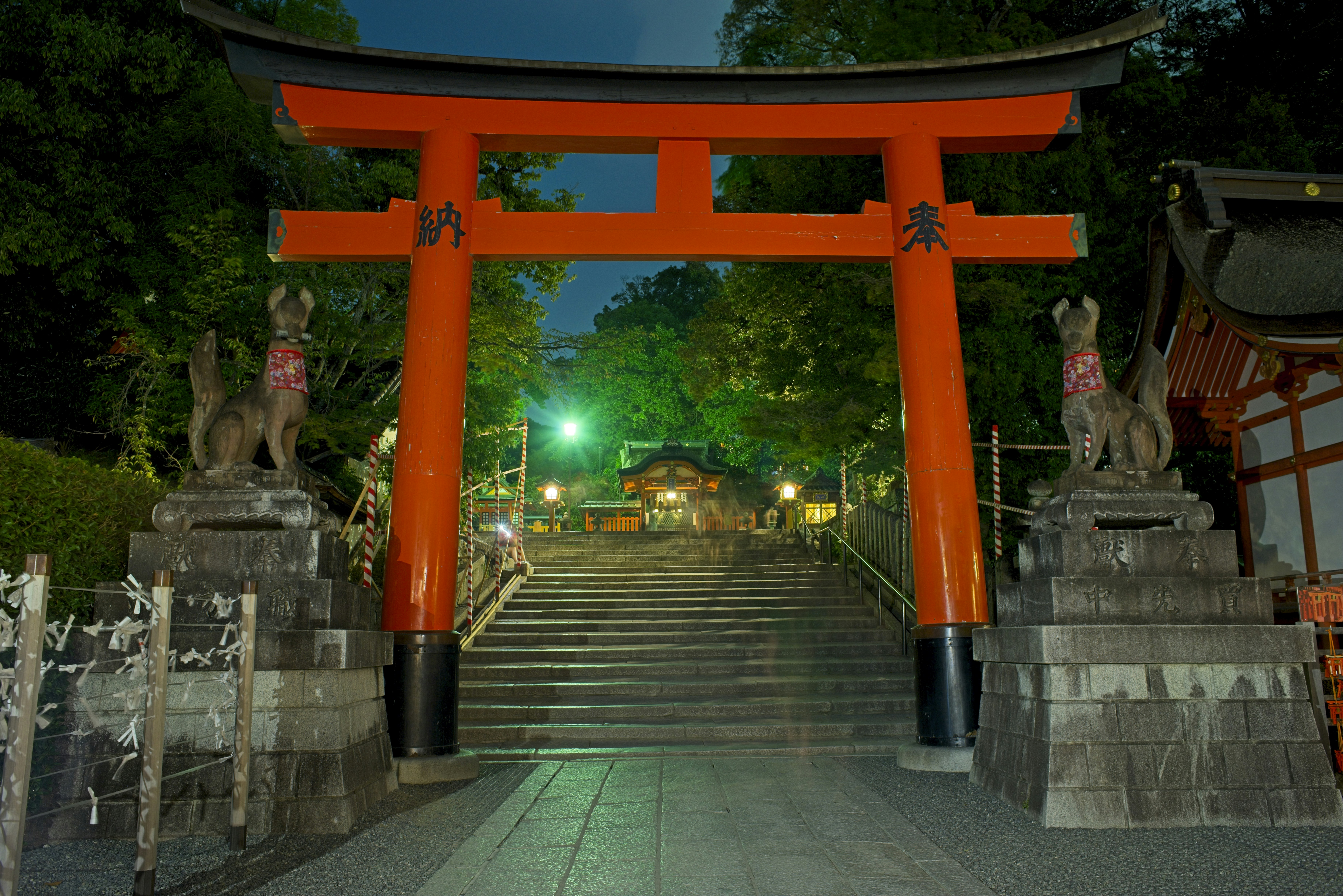 A long exposure captures a grand vermillion torii gate flanked by weathered stone kitsune guardian statues marks the entrance stairway to Fushimi Inari Taisha's main shrine hall, Kyoto, at dusk on a humid July evening. Illuminated by warm lantern light and a green-tinged spotlight, the stone steps lead upward toward the ornate shrine building nestled among lush trees beneath a deepening twilight sky, capturing the sacred approach to this iconic Kyoto landmark.