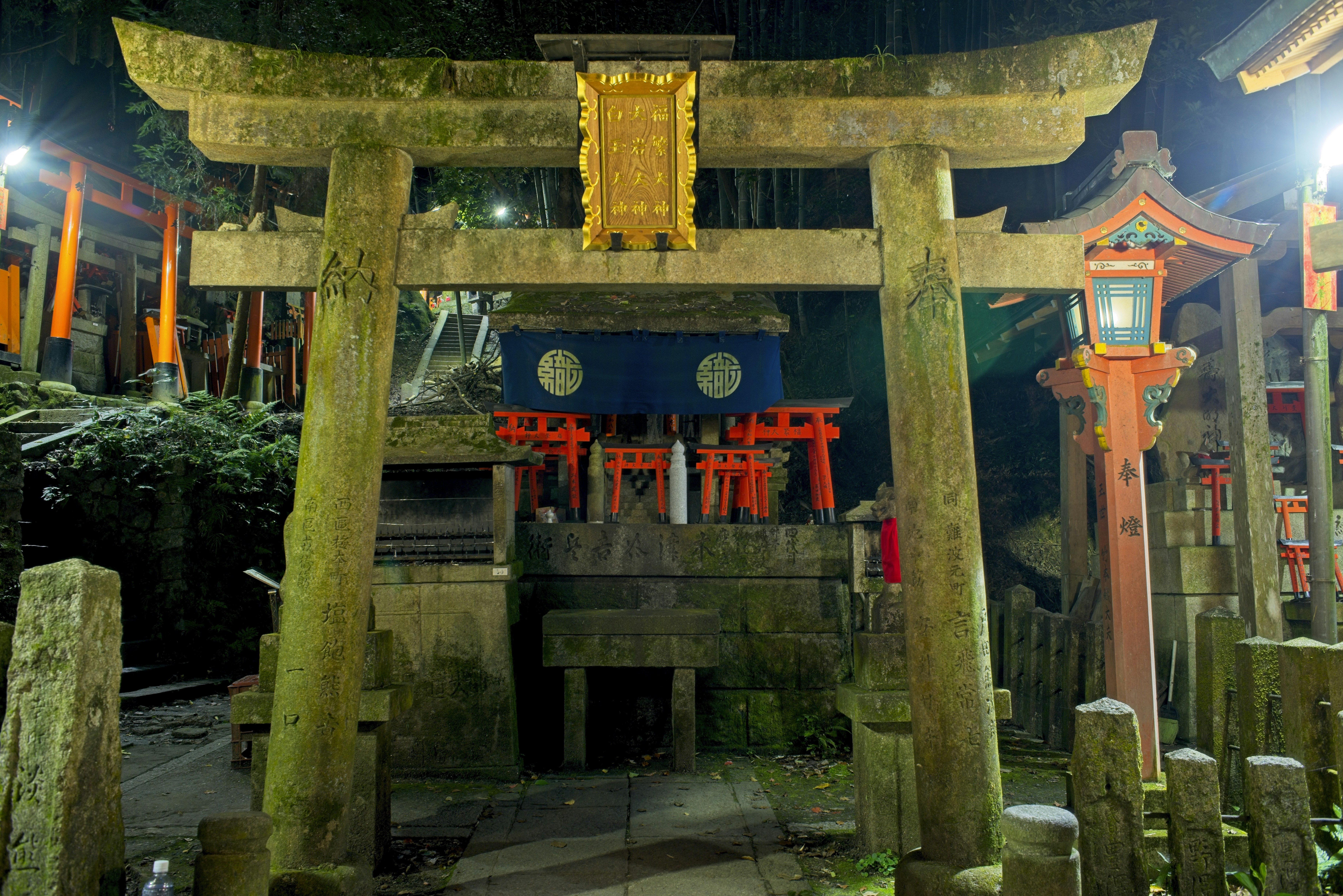 A long exposure captures a moss-covered stone torii gate frames a small shrine altar adorned with a golden inscribed plaque and blue noren curtain at Fushimi Inari Taisha's mountain trail, Kyoto, on a humid July evening. Miniature orange torii gates cluster around a central stone monument, while an illuminated lantern glows softly to the right. Weathered stone pillars bearing carved kanji lead deeper into this atmospheric, spiritually significant corner of the sacred mountain path.