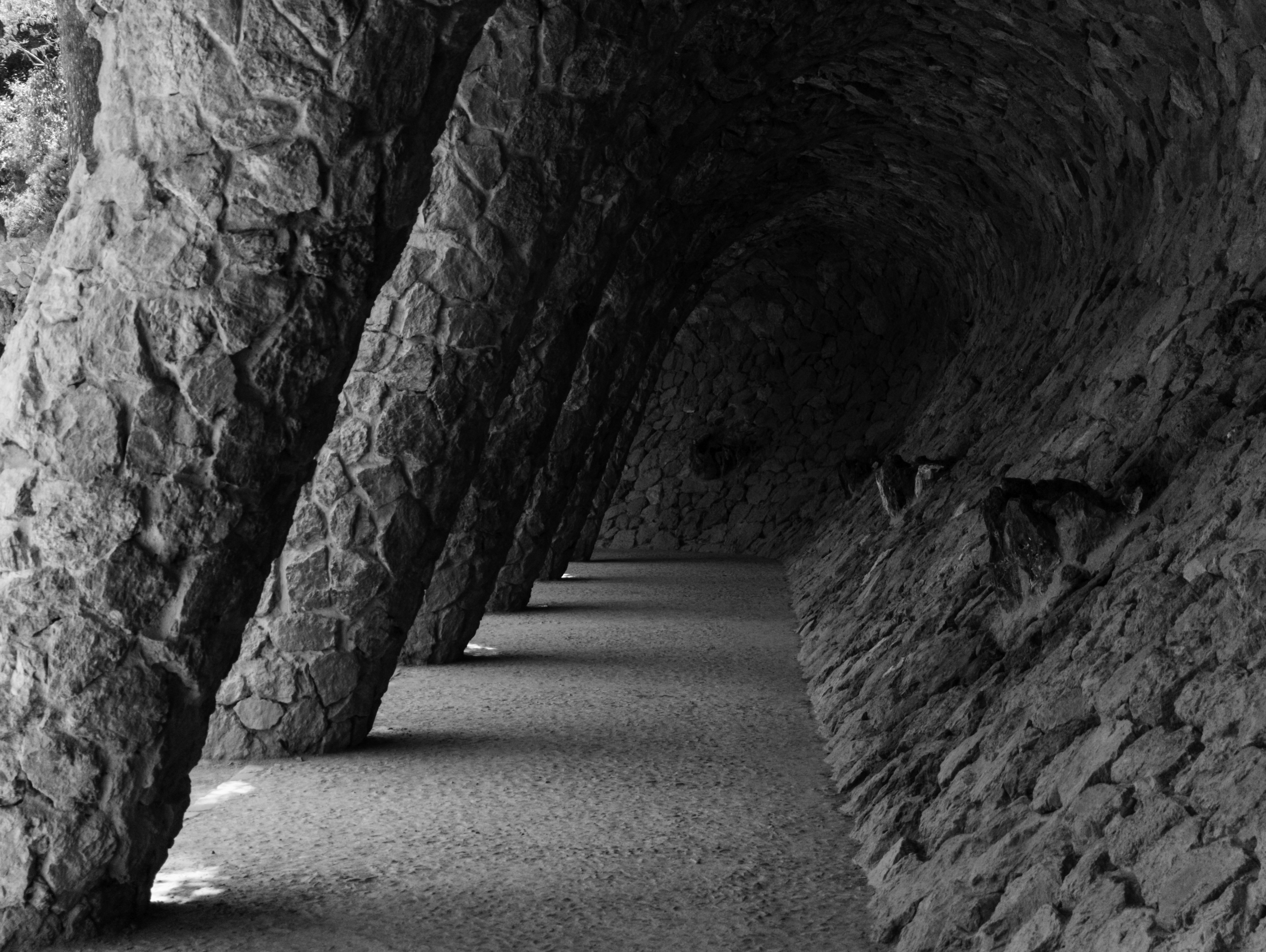 Curved stone archway forms a long, dim corridor with textured rock walls and a pebbled floor. The scene emphasizes depth and repetition in grayscale, suggesting an ancient or tunnel-like passage.