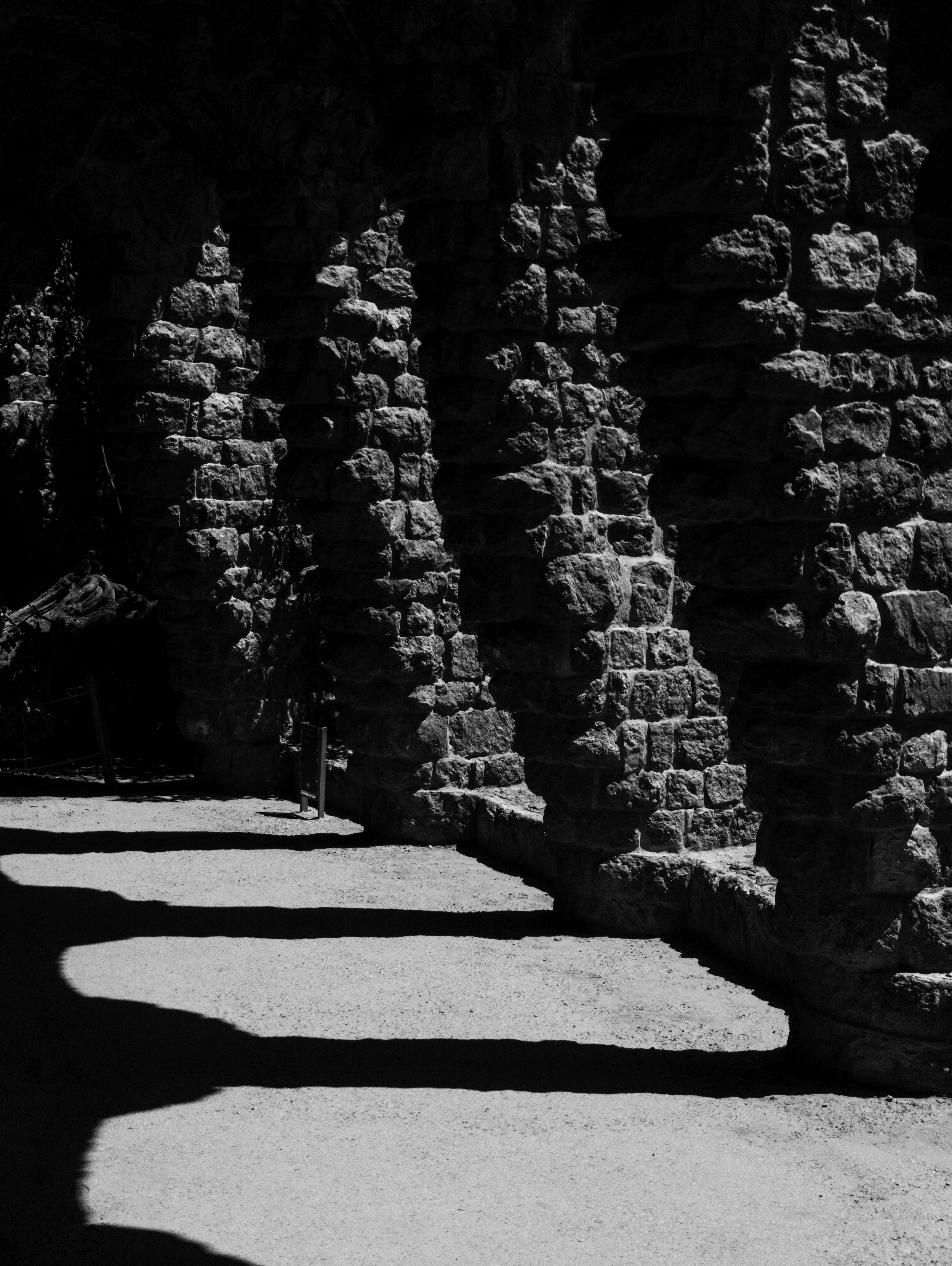 High-contrast black-and-white view of sunlit stone arches casting deep shadows along a walkway, emphasizing texture and geometry.