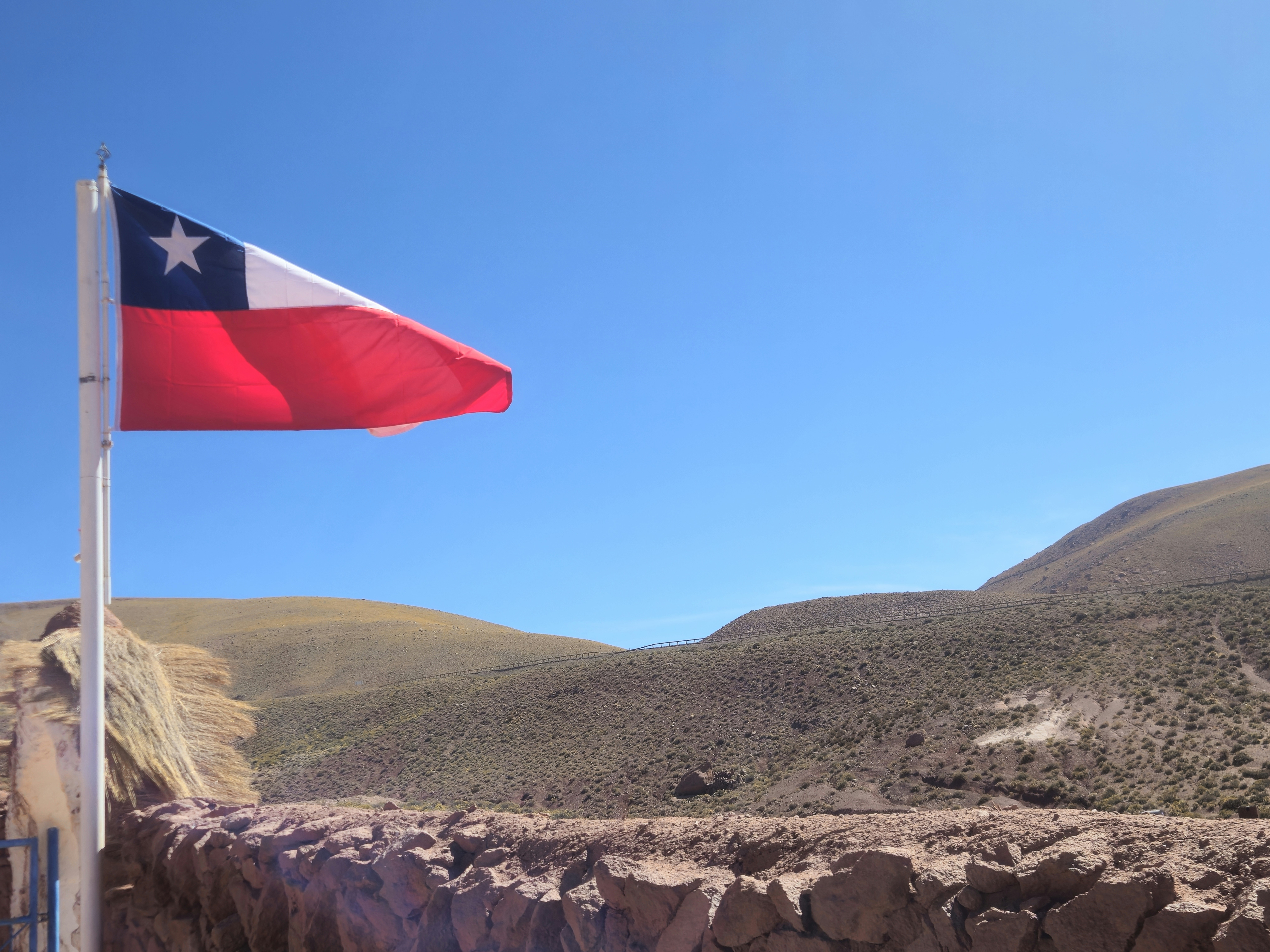 The flag of Chile waving on a pole above a stone wall and arid hills