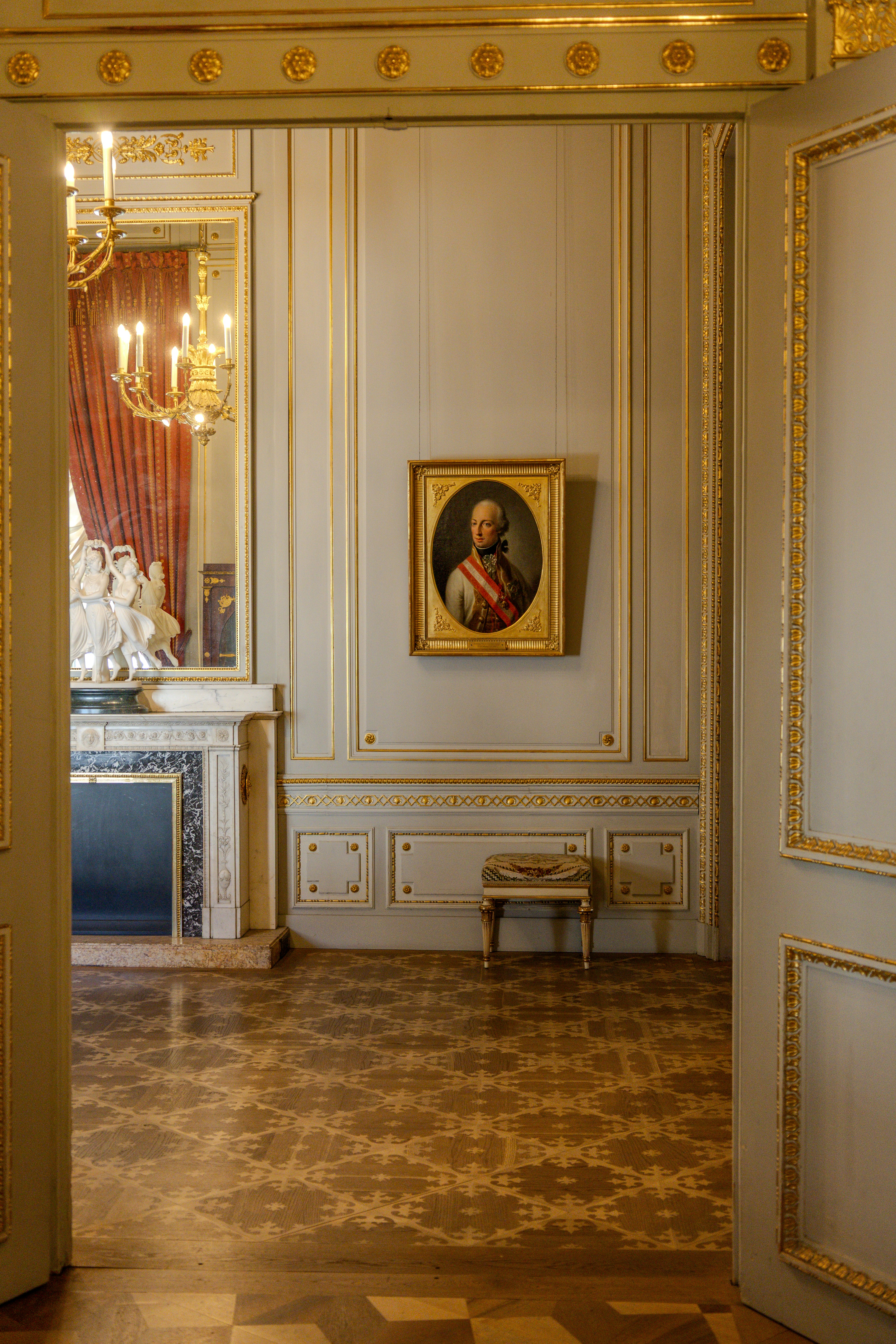 Elegant European parlor with pale walls, gold trim, and a central portrait in a gilded frame; chandelier reflections and a marble mantel hint at opulent interior design.