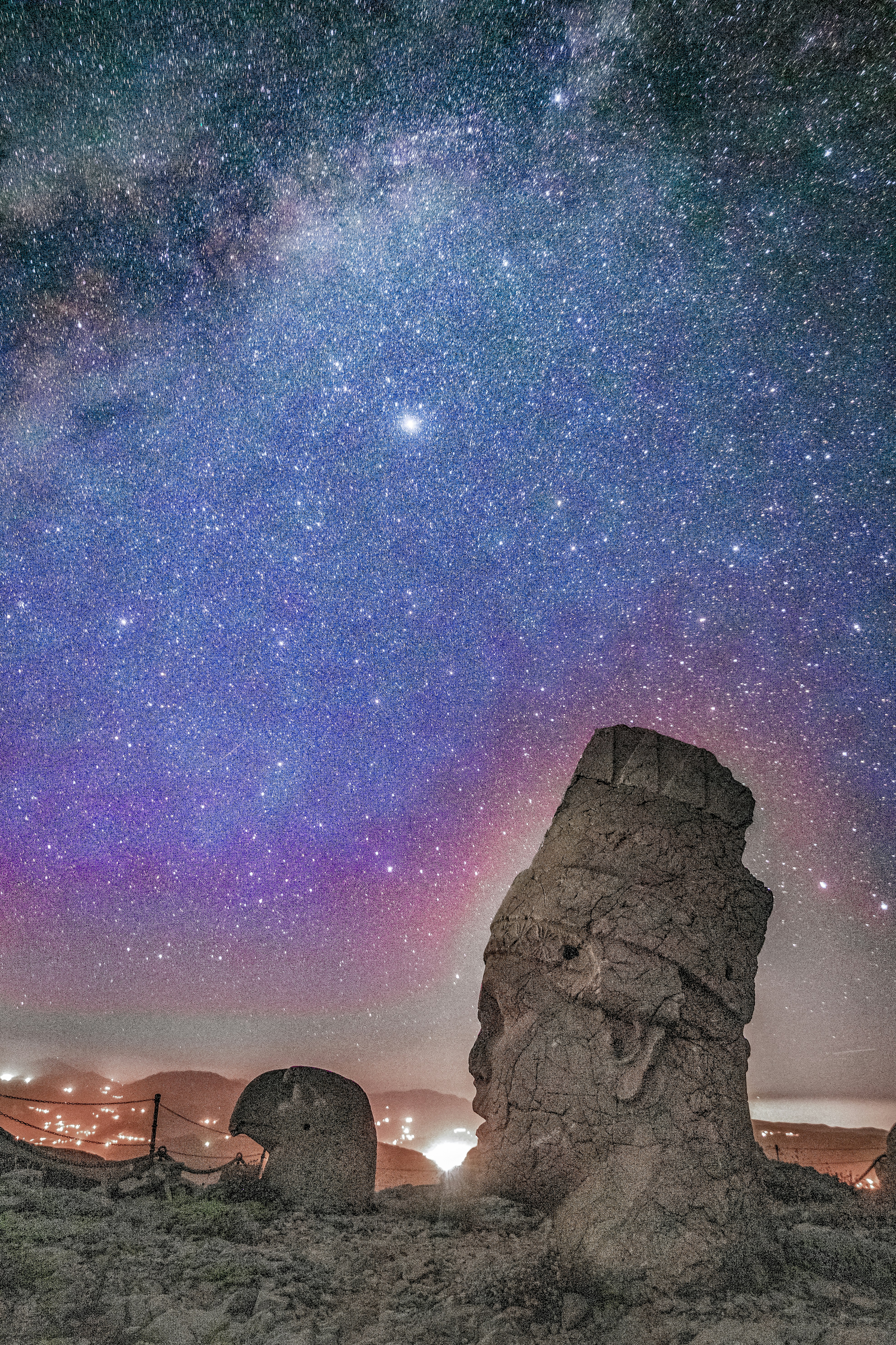 A rugged rock formation rises beside a smaller stone sculpture as a colorful Milky Way arches overhead, with distant lights along the horizon.