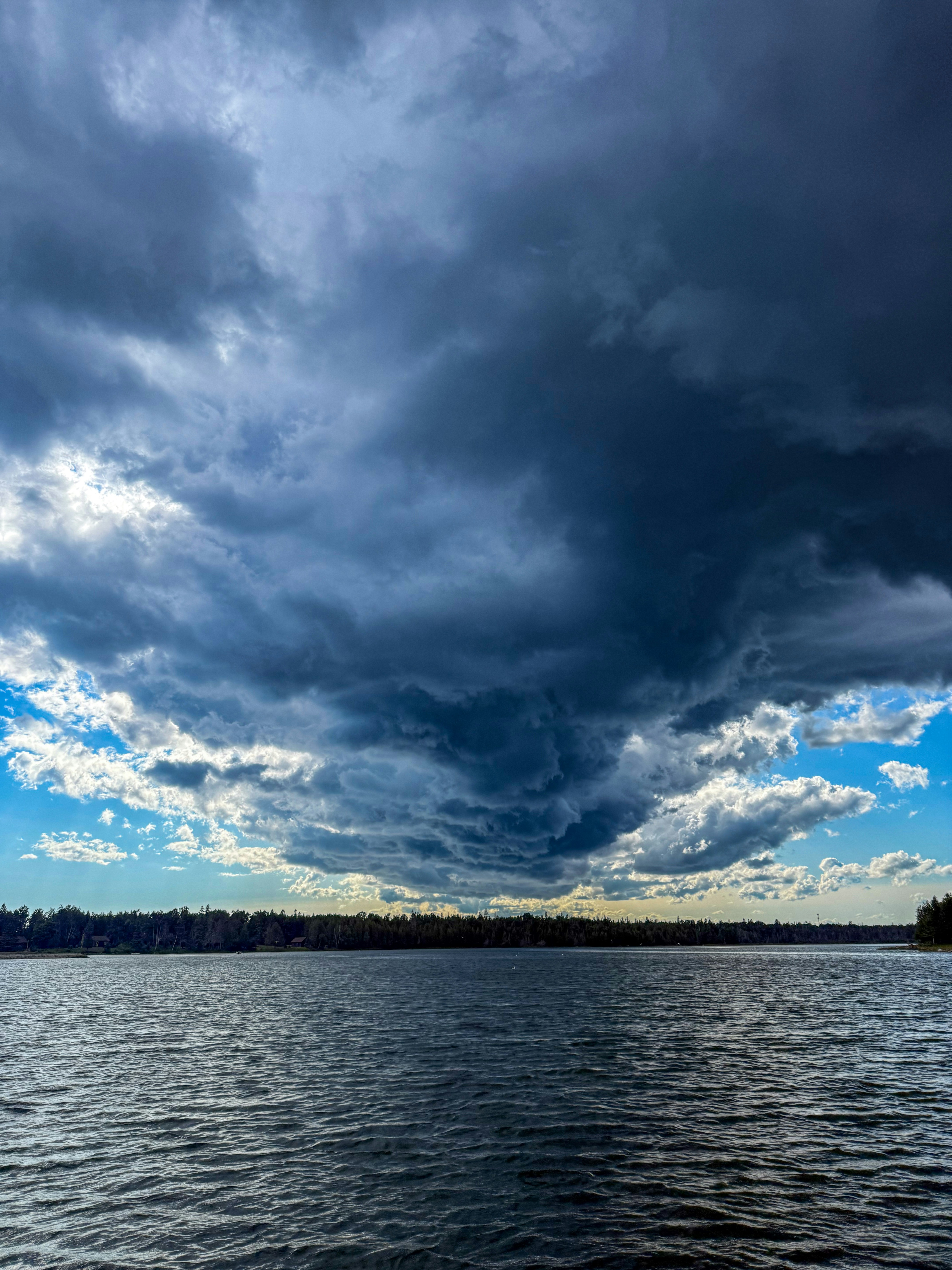 A tranquil lake stretches to a dark, dramatic sky; distant tree line anchors the horizon, emphasizing the contrast between calm water and turbulent clouds. This is a photographic capture of natural scenery.