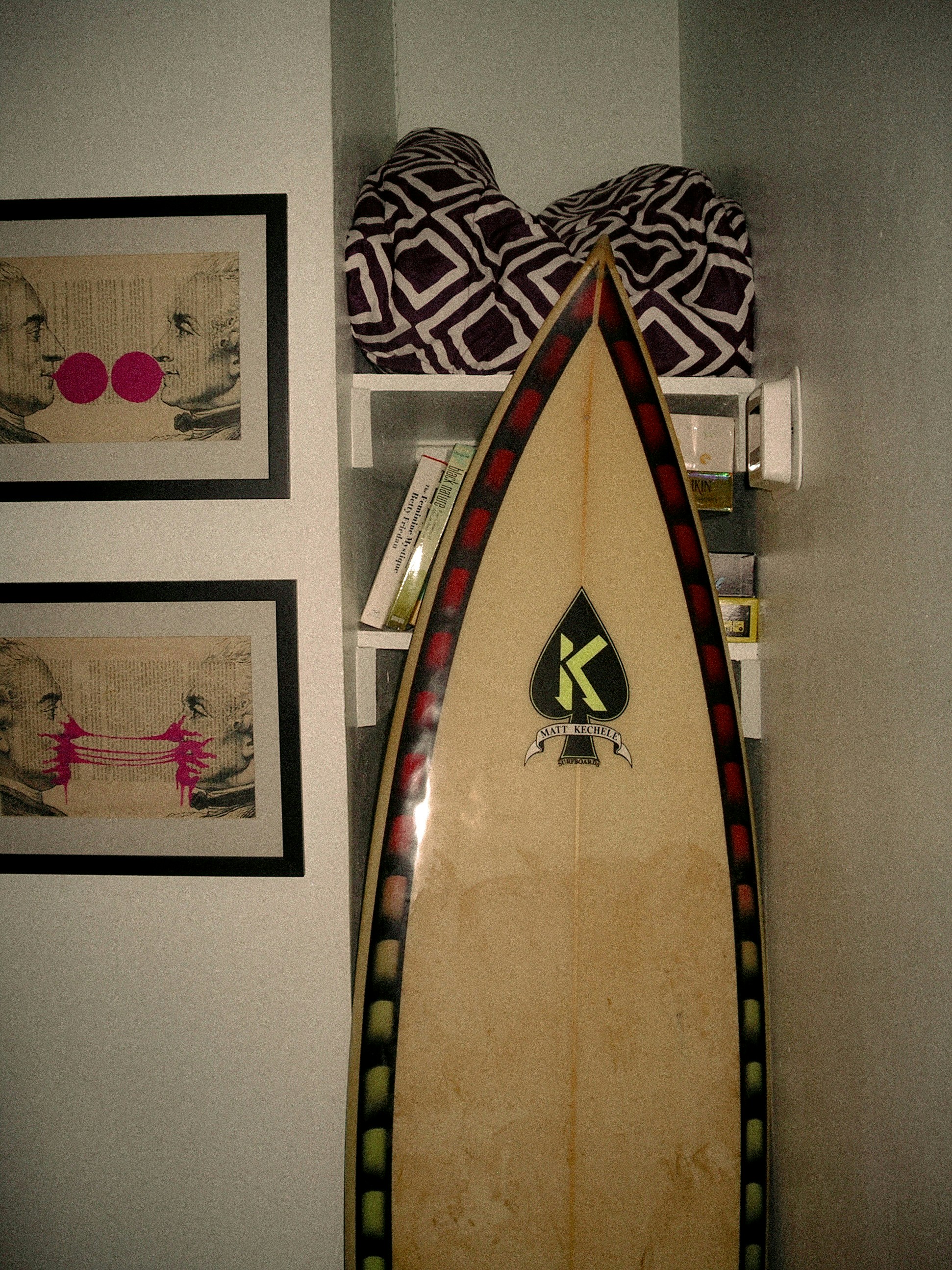 A surfboard leaning in a corner beside framed prints and a patterned cushion.